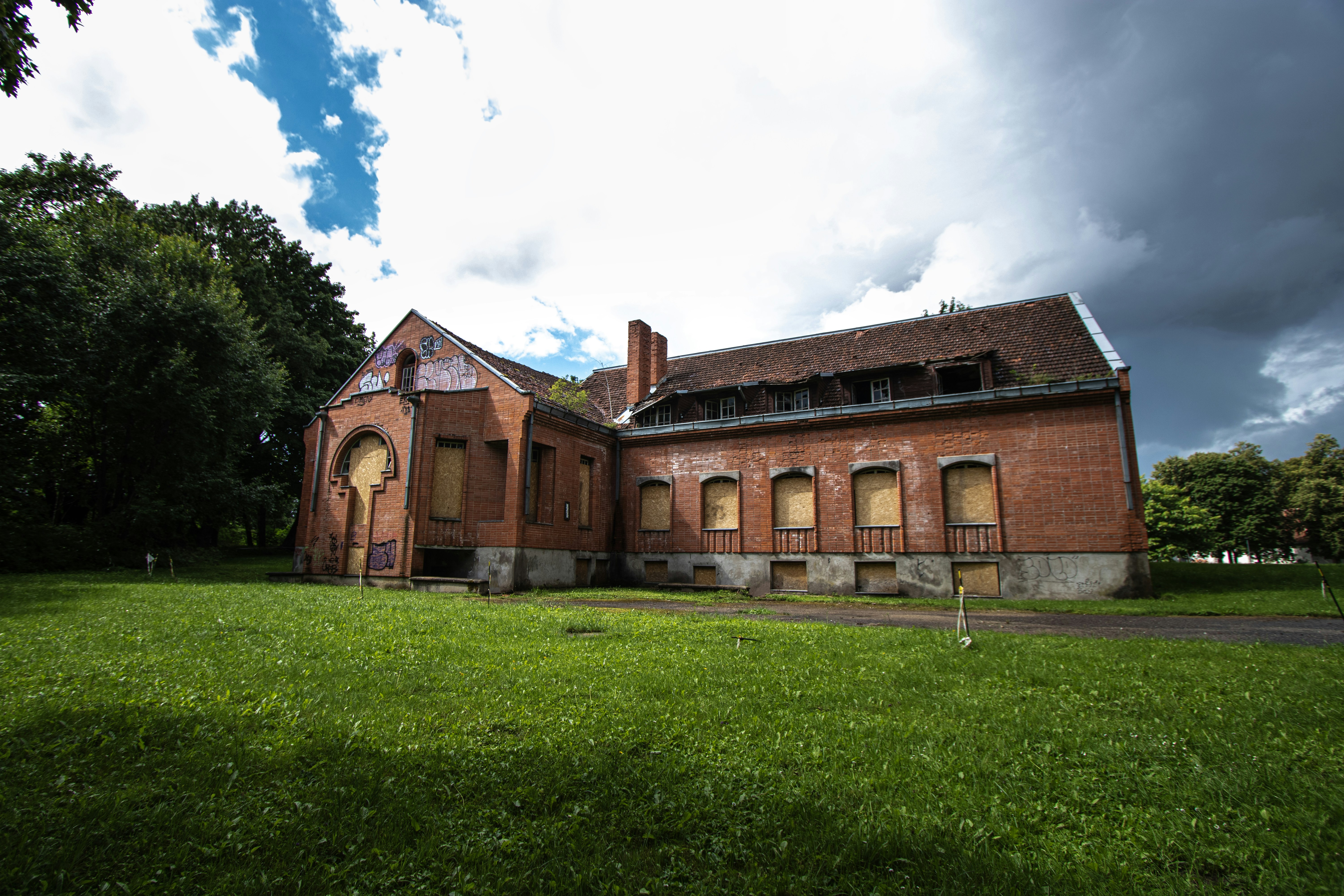 A brick building with arched doorway sits in a grassy clearing, under a mix of blue sky and dark clouds.