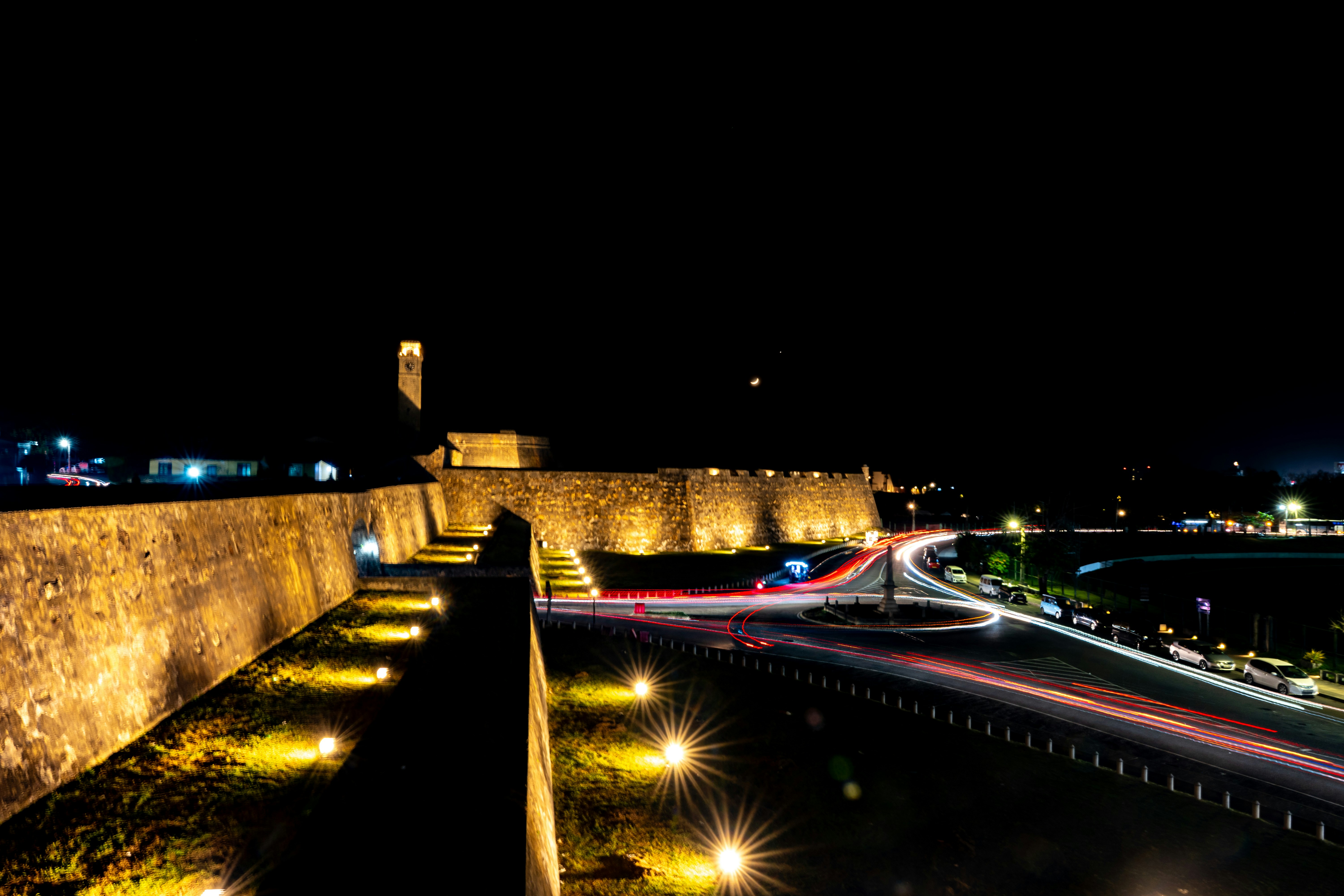 Long-exposure shot of a historic fortress wall and roadway at night, with light trails along a curved bridge and warm illumination on the stonework.