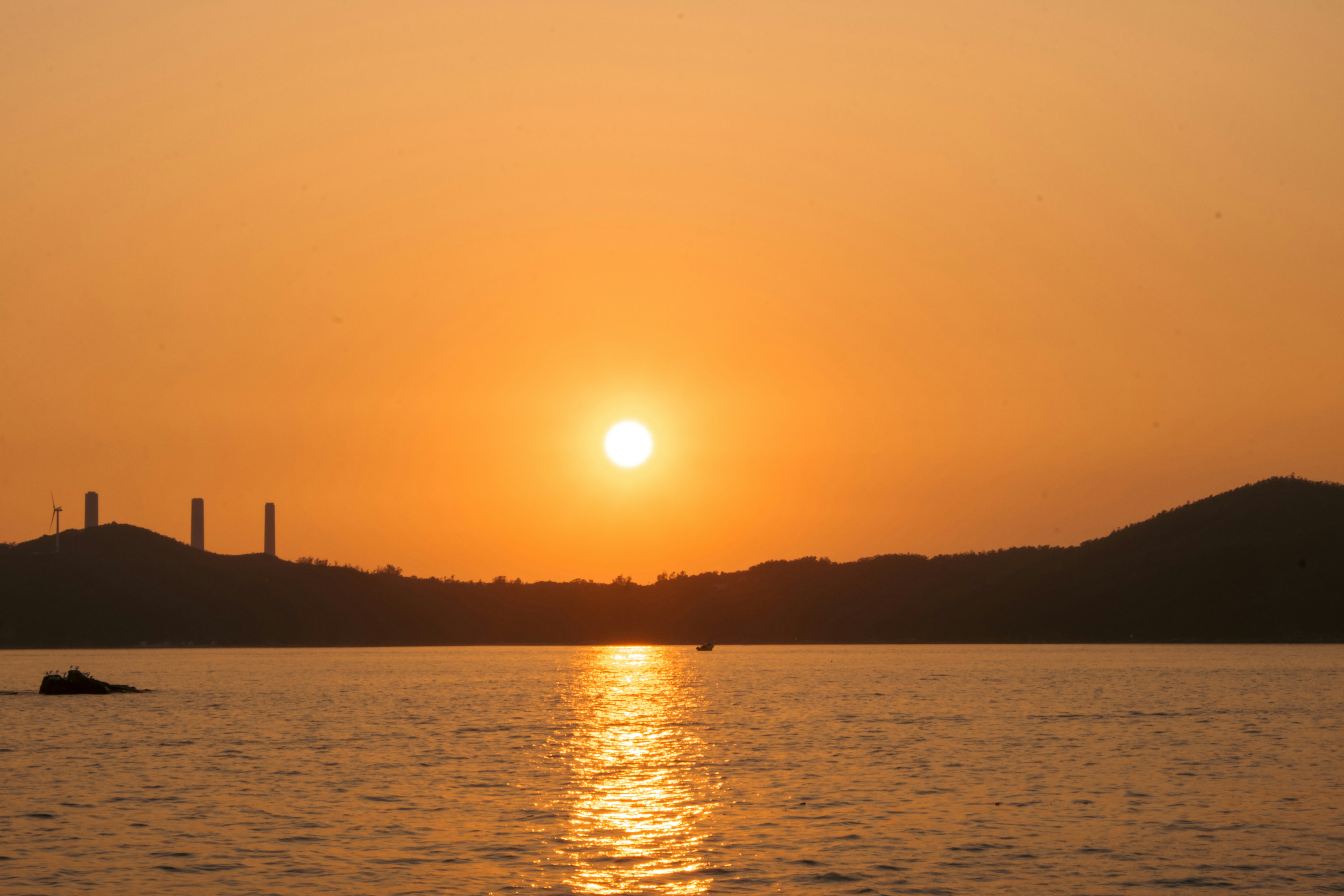 A beautiful orange golden sunset over the sea off Ap Lei Chau, Hong Kong, casting a shimmering light reflection across the water toward the silhouetted hills and distinct power station chimneys of Lamma Island.