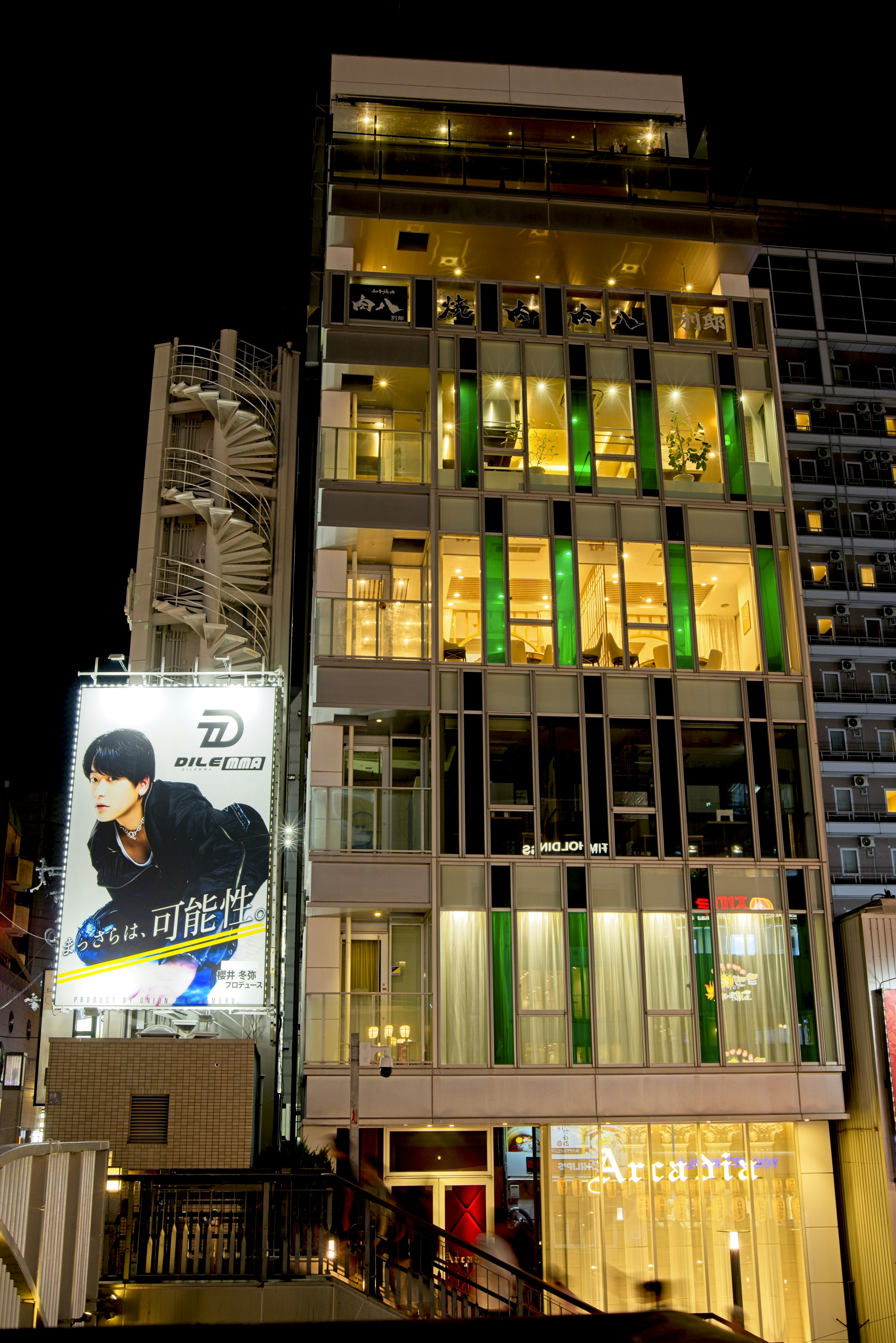 A tall, brightly lit building near Dotonbori in Osaka showcases layers of glowing green and gold-lit windows, revealing cozy restaurant interiors within, on a hot summer evening in July. A large billboard featuring a Japanese celebrity advertises a fashion or cosmetics brand along the building's side, illuminated with bright LED trim. A striking spiral staircase adds architectural interest above, while the ground floor entrance glows with warm light beneath an "Arcadia" sign. This vertical, illuminated facade captures the layered visual density typical of Osaka's nighttime cityscape.