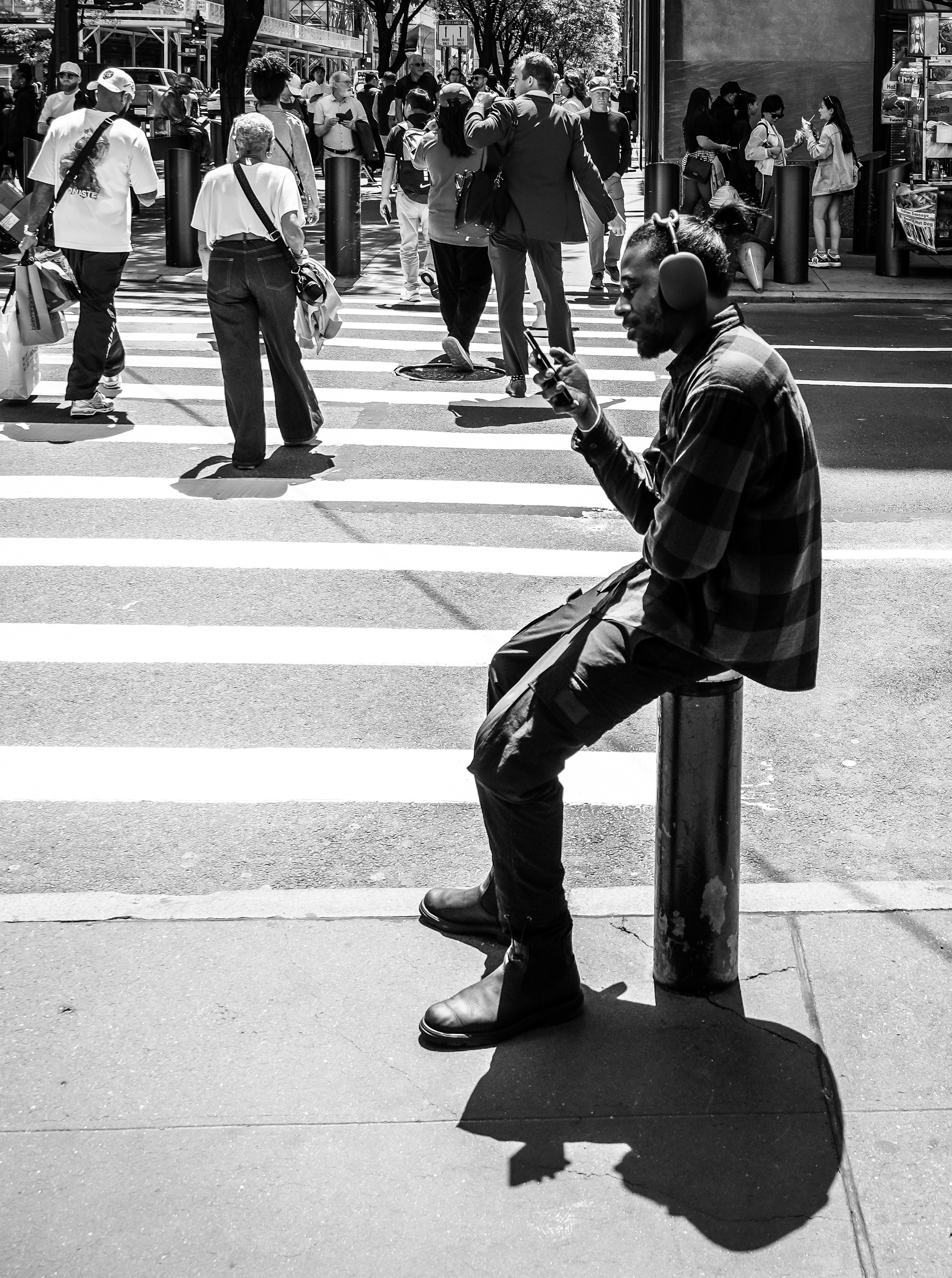 A solitary man sits on a bollard at a busy crosswalk, headphones on and phone in hand, while pedestrians pass by in the background on a sunny city street.