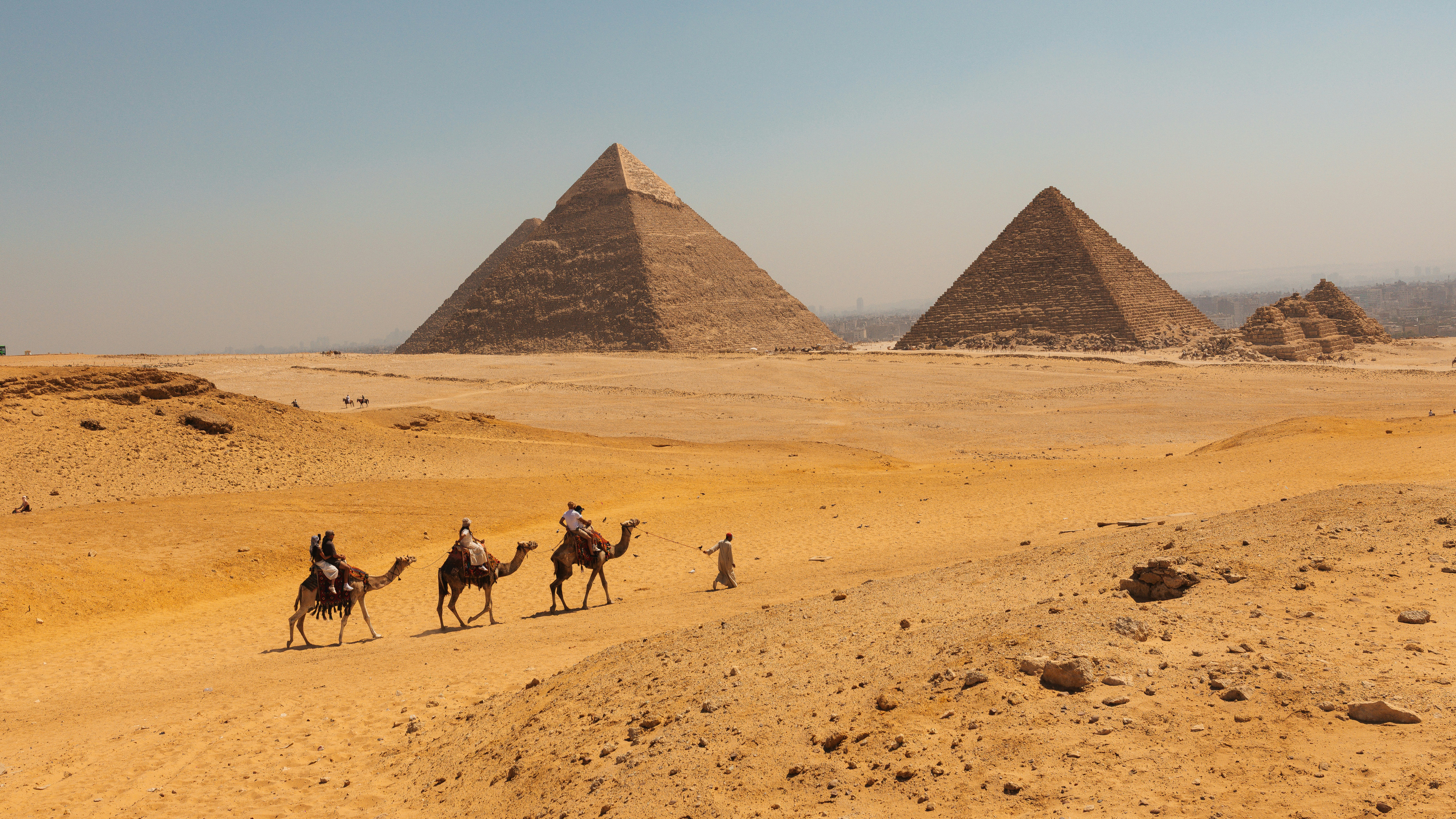 Caravan drift across the desert toward the pyramids under a pale sky