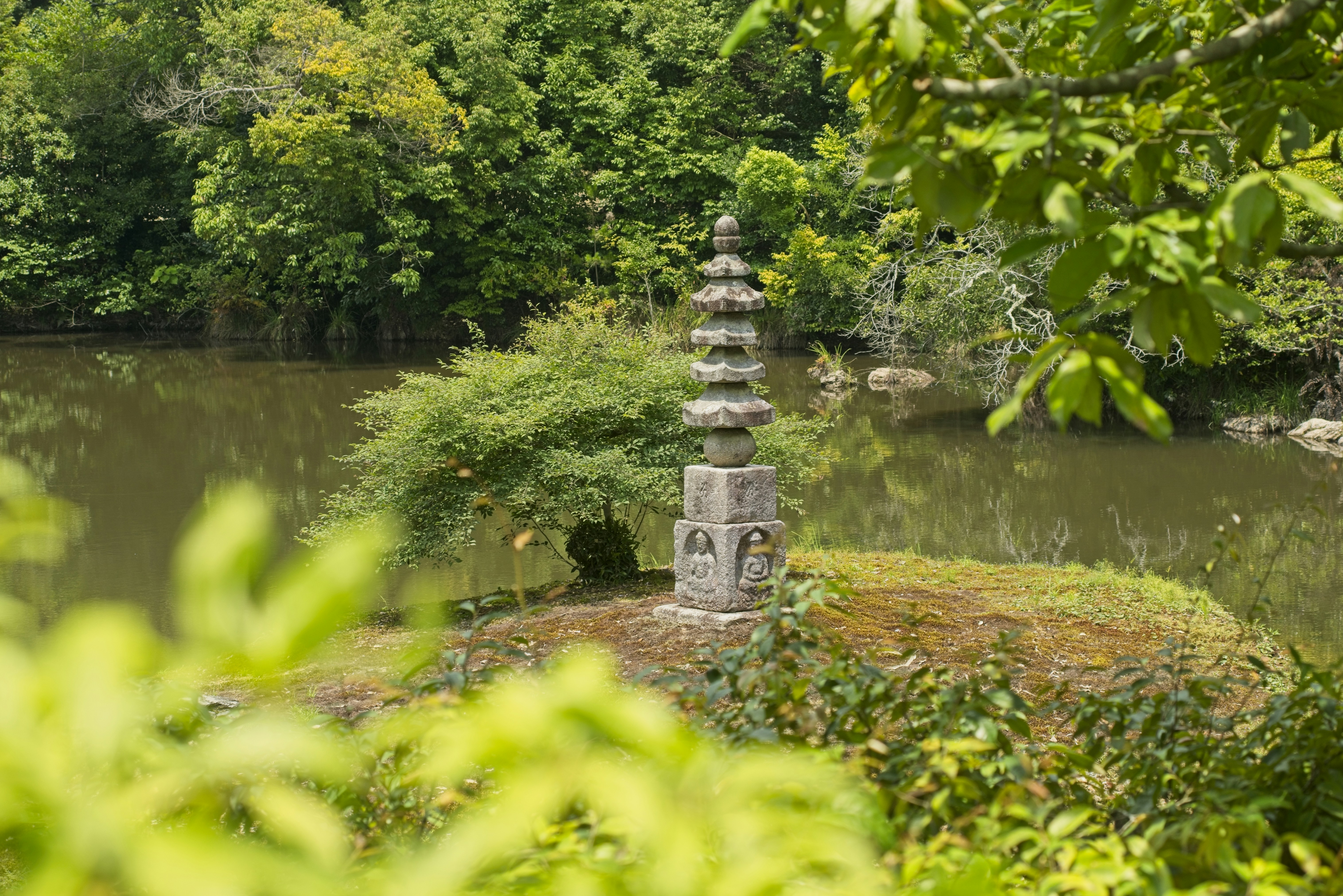A small stone five-tiered pagoda stands quietly on a tiny island within a tranquil pond near Kinkaku-ji in Kyoto, framed naturally by sunlit green foliage in the foreground on a hot summer day at noon in July. Dense forest and shrubbery line the calm water's edge, their reflections shimmering gently across the surface. The weathered stone tower, adorned with subtle carvings, adds a serene focal point to the lush garden setting. This peaceful, secluded corner exemplifies the harmonious blend of nature and traditional craftsmanship found throughout Kyoto's temple gardens.