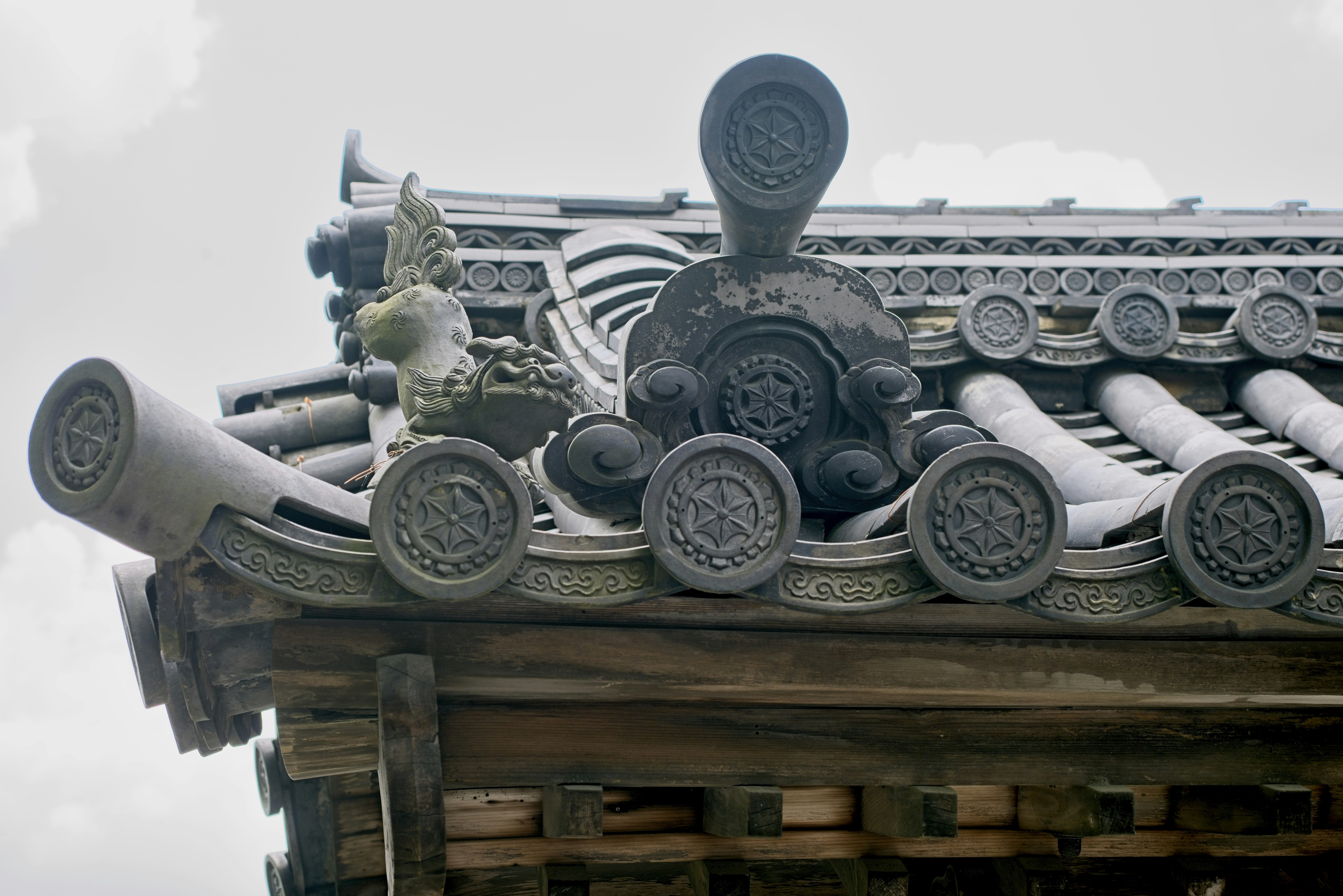 A close-up view of traditional Japanese roof tiles showcases the intricate craftsmanship of temple architecture near Kinkaku-ji in Kyoto on a hot summer day at noon in July. Ornate circular end-caps decorated with floral motifs line the curved eaves, while a small mythical creature figure perches near the roof's edge. The dark, weathered ceramic tiles contrast against the pale overcast sky, highlighting the fine detail and symmetry of the design. This architectural close-up reflects the meticulous artistry found throughout Kyoto's historic temple structures.