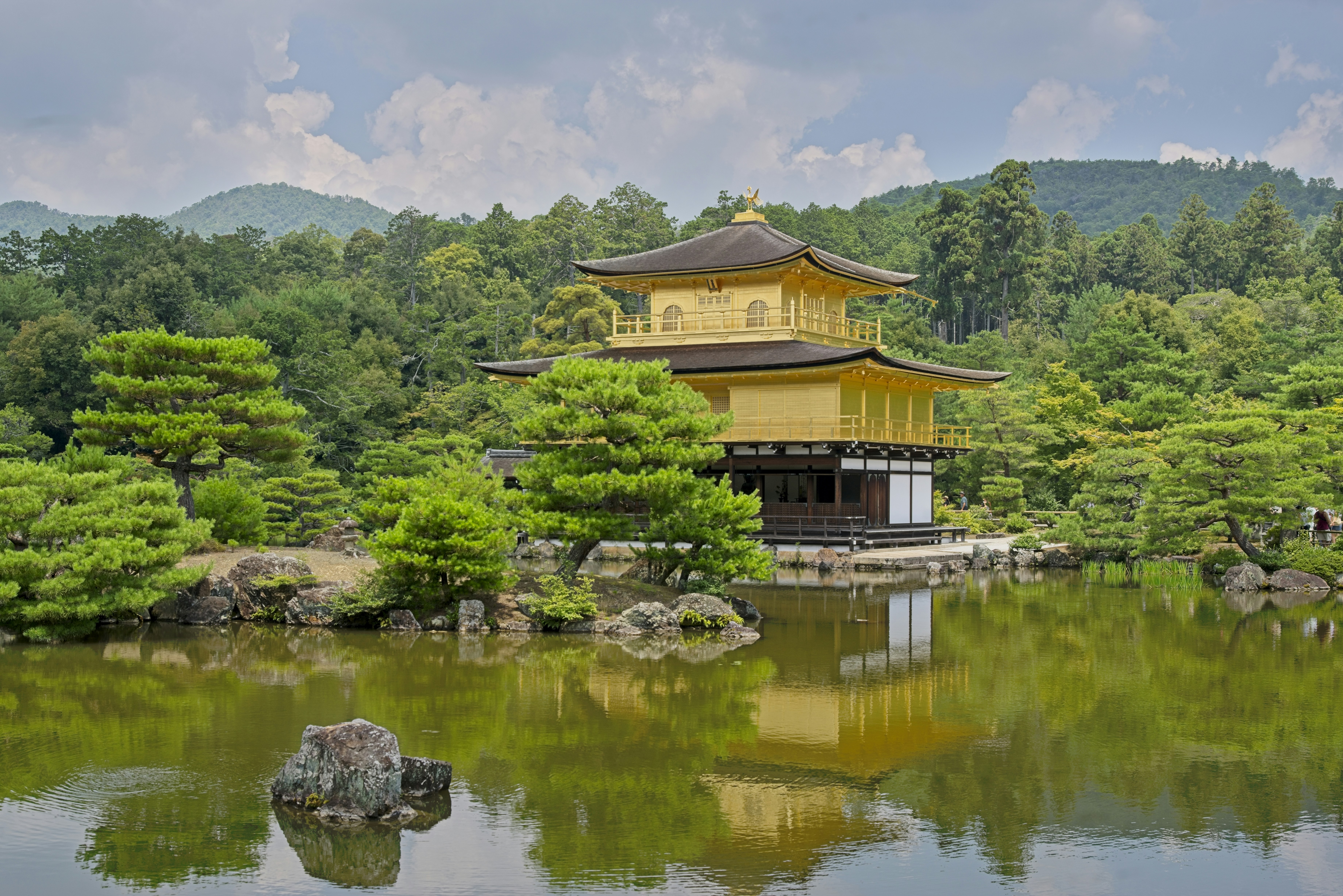 The dazzling Golden Pavilion of Kinkaku-ji rises above its tranquil reflecting pond in Kyoto, its gold-leafed exterior gleaming beautifully against a backdrop of dense green forest and distant mountains on a hot summer day at noon in July. A golden phoenix ornament crowns the layered rooftop, while carefully pruned pine trees and scattered rocks frame the shoreline. The temple's mirror image shimmers gently across the still water, creating a striking symmetrical composition. This iconic Zen Buddhist temple remains one of Japan's most celebrated and photographed landmarks.