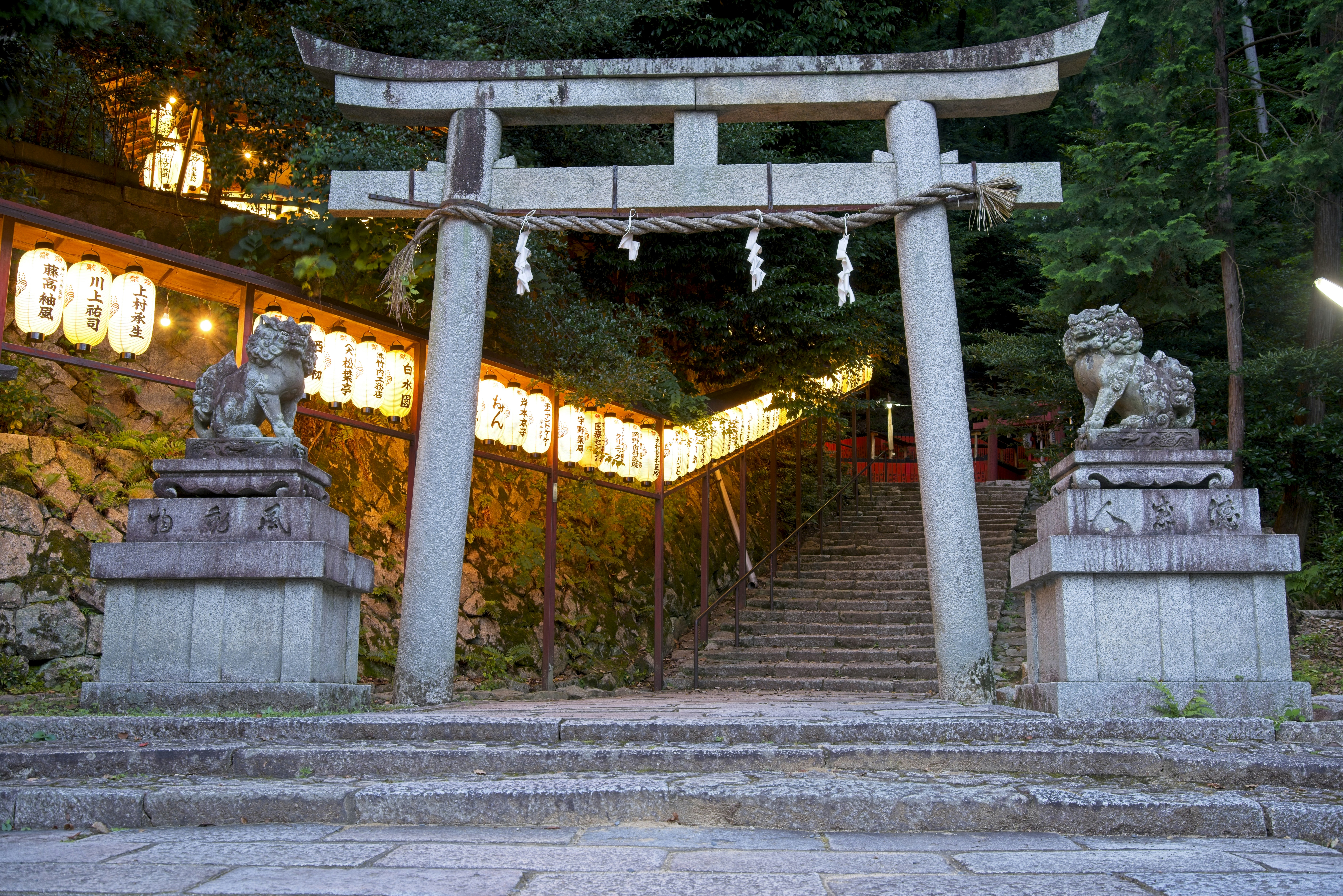 A weathered stone torii gate marks the entrance to a shrine stairway in Kyoto, flanked by two guardian komainu lion statues on stone pedestals, as glowing paper lanterns illuminate the ascending steps at dusk. A thick sacred shimenawa rope with hanging paper streamers hangs across the gate's crossbeam, while a row of lanterns bearing donor names lines the stairway railing, leading up toward a red-lit structure in the distance. Dense forest frames the scene on both sides. This atmospheric approach captures the quiet reverence and traditional craftsmanship found at Kyoto's shrine sites.