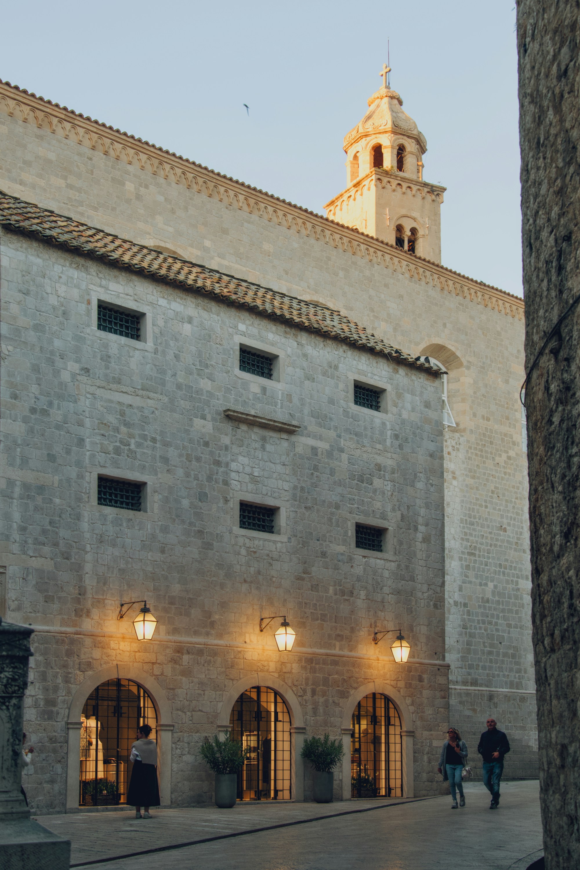 People walking past a stone building with arched doorways and glowing lanterns in Dubrovnik