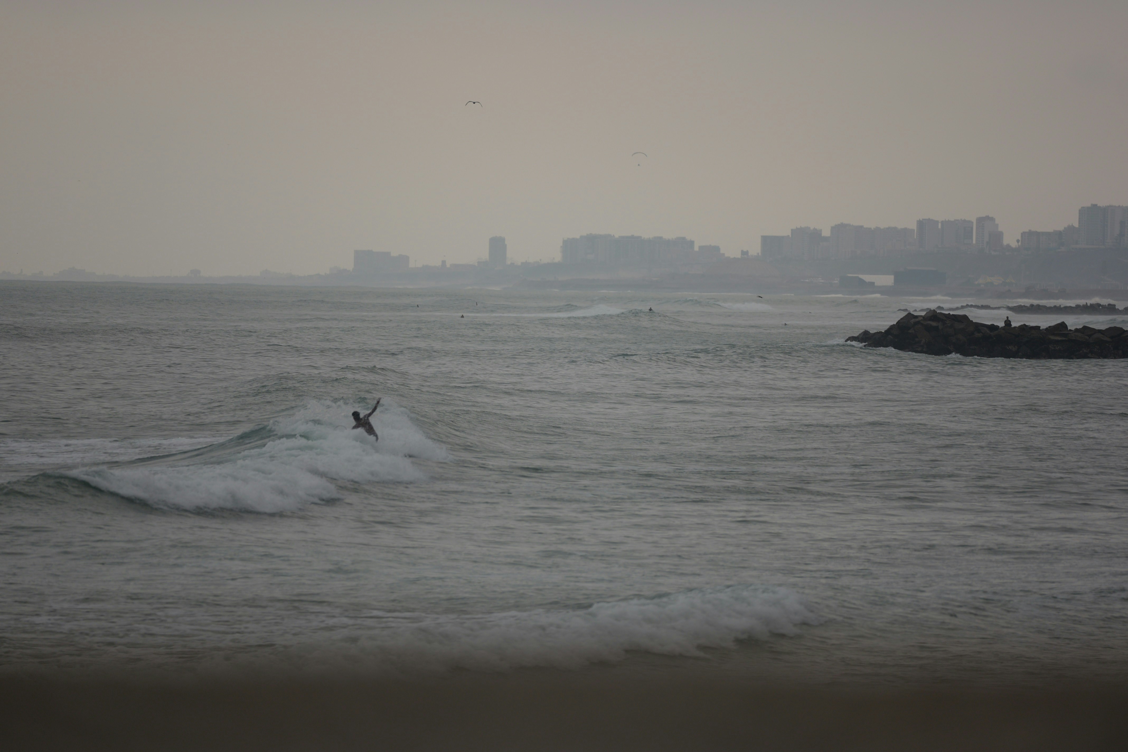 Surfer in a Pale Coastal Wake under Distant City Veil