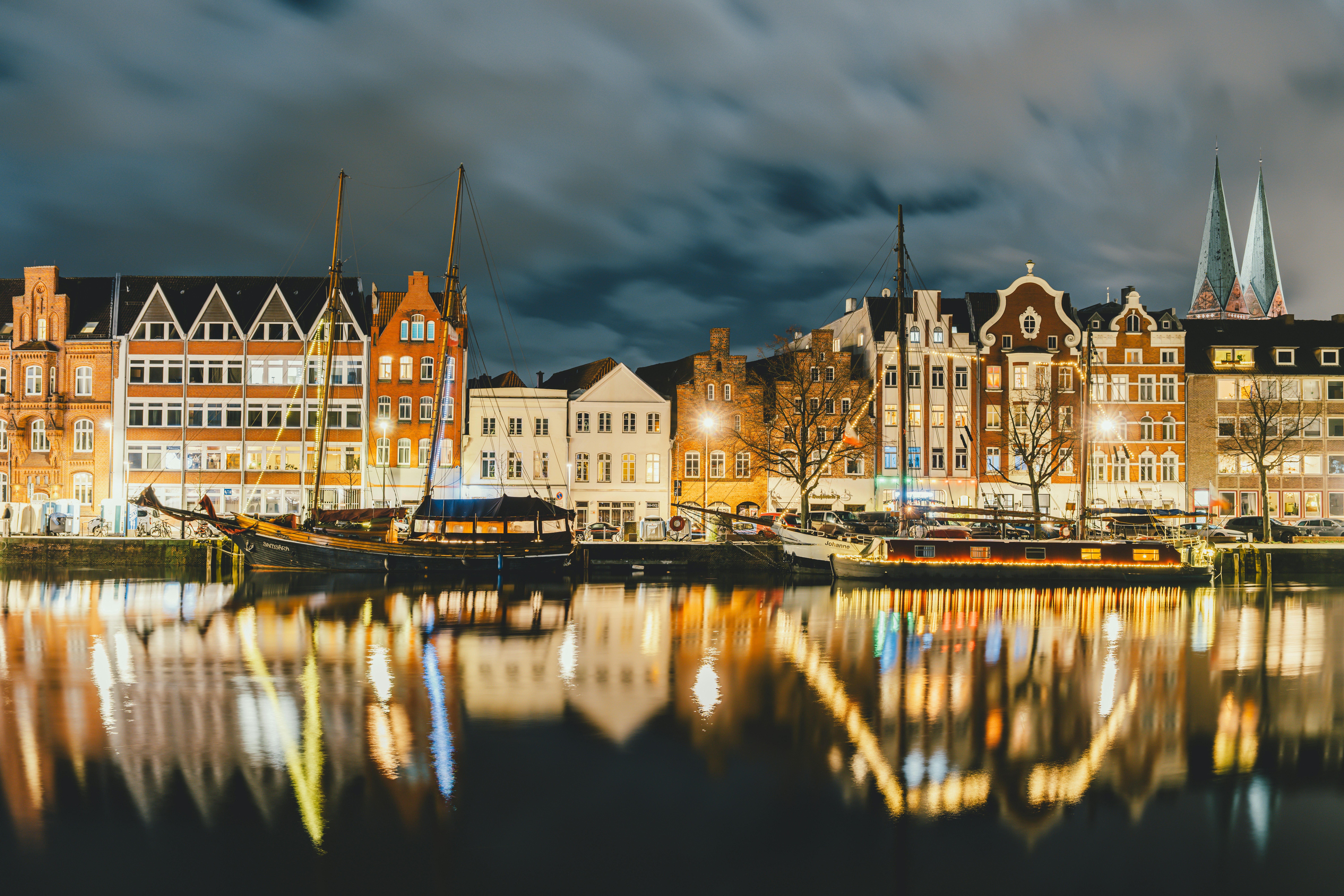 Night harbor scene with illuminated historic buildings and their reflections on calm water; boats docked along the quay, a moody sky overhead. A cityscape photograph capturing warm tones and mirrored architecture.