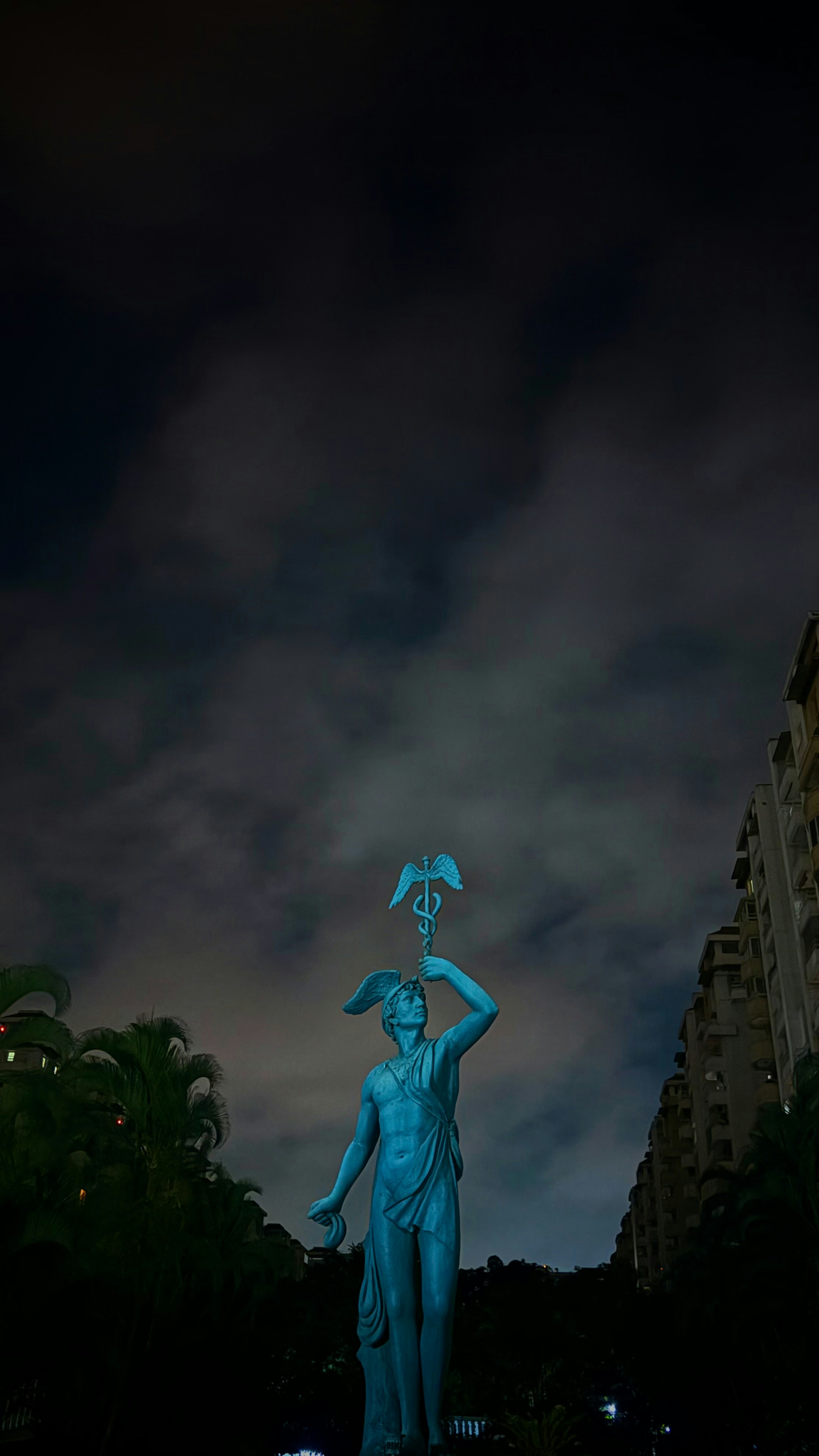 Blue statue illuminated at night with surrounding buildings and dark sky, highlighting a monument in a park setting.