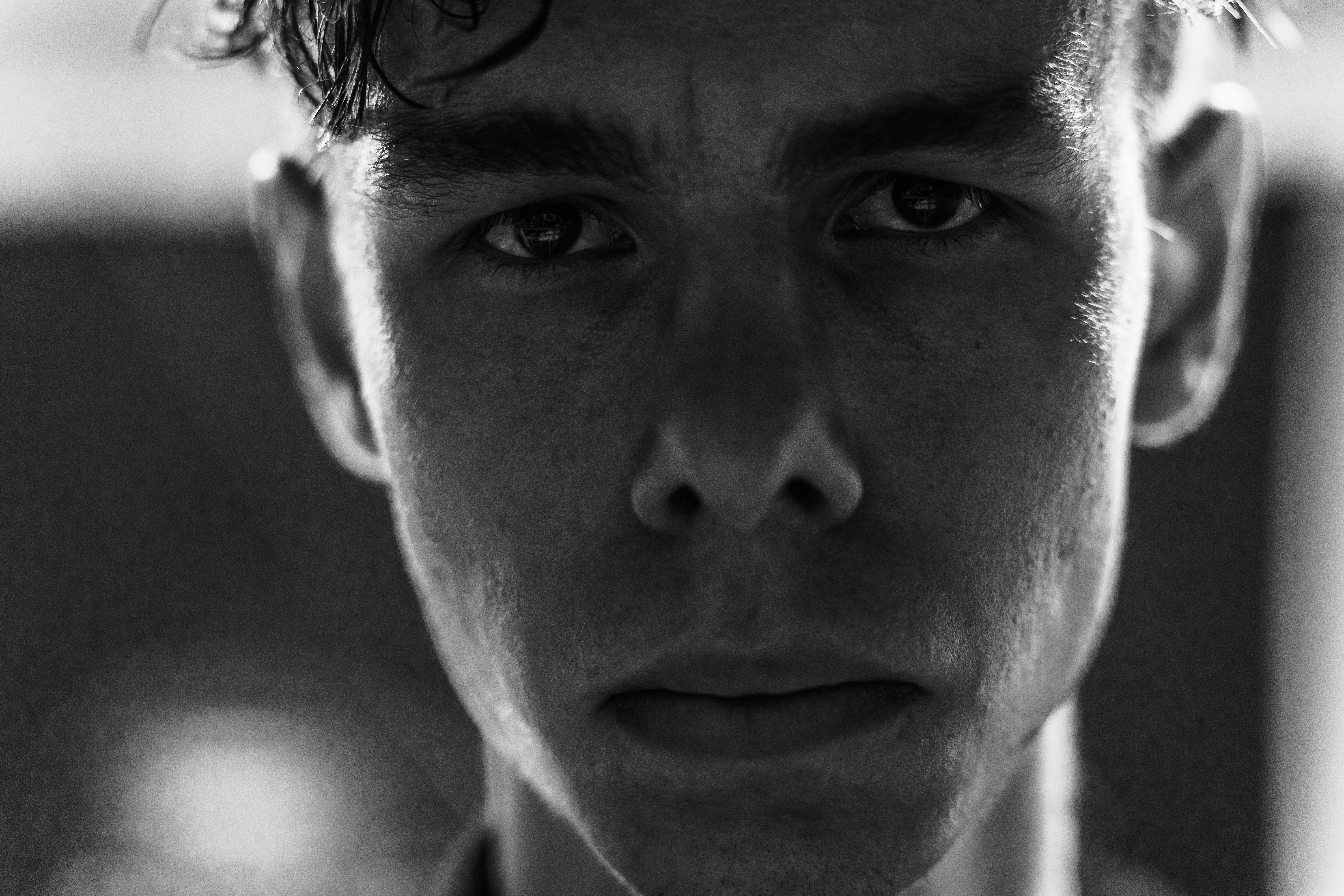 Intense close-up portrait highlighting the subject's eyes and serious expression in black and white. A stark, square crop emphasizes texture and mood without background distraction.