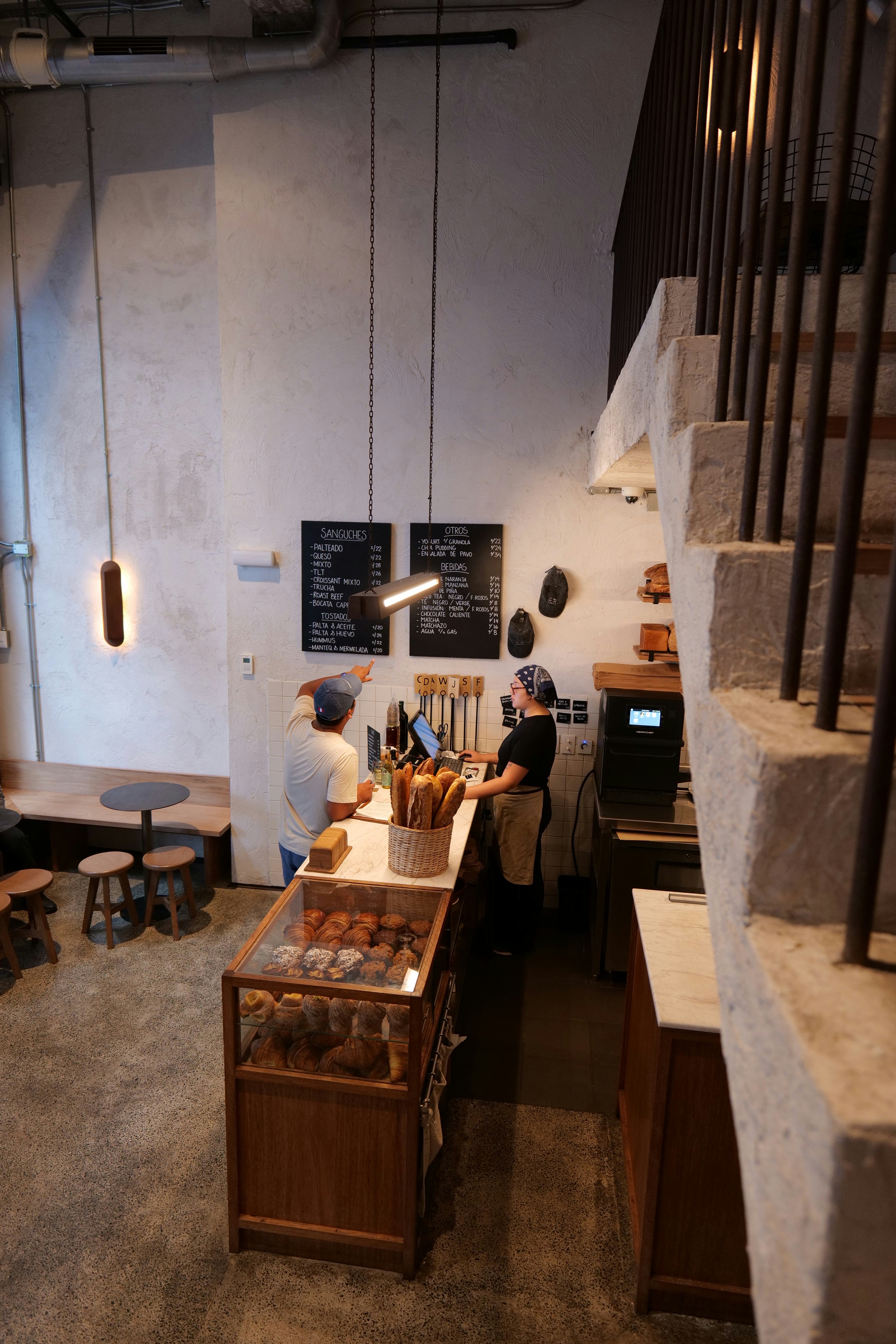 Warm cafe scene with two baristas behind a pastry counter in a minimalist, industrial space; soft lighting and concrete walls emphasize the counterwork and baked goods.