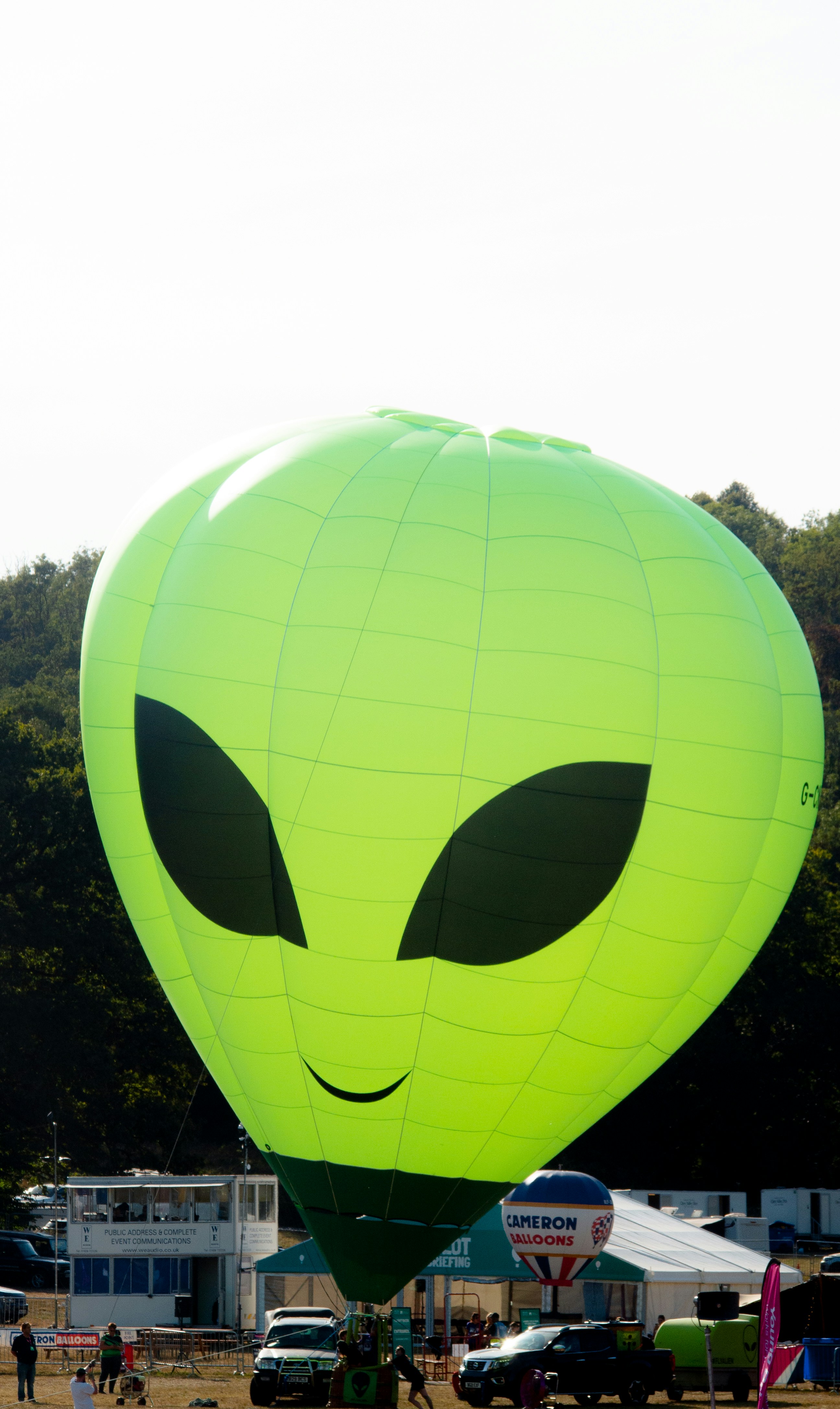 Bright lime-green alien-head balloon towers over festival tents and vehicles, serving as the focal attraction in an outdoor fair scene.