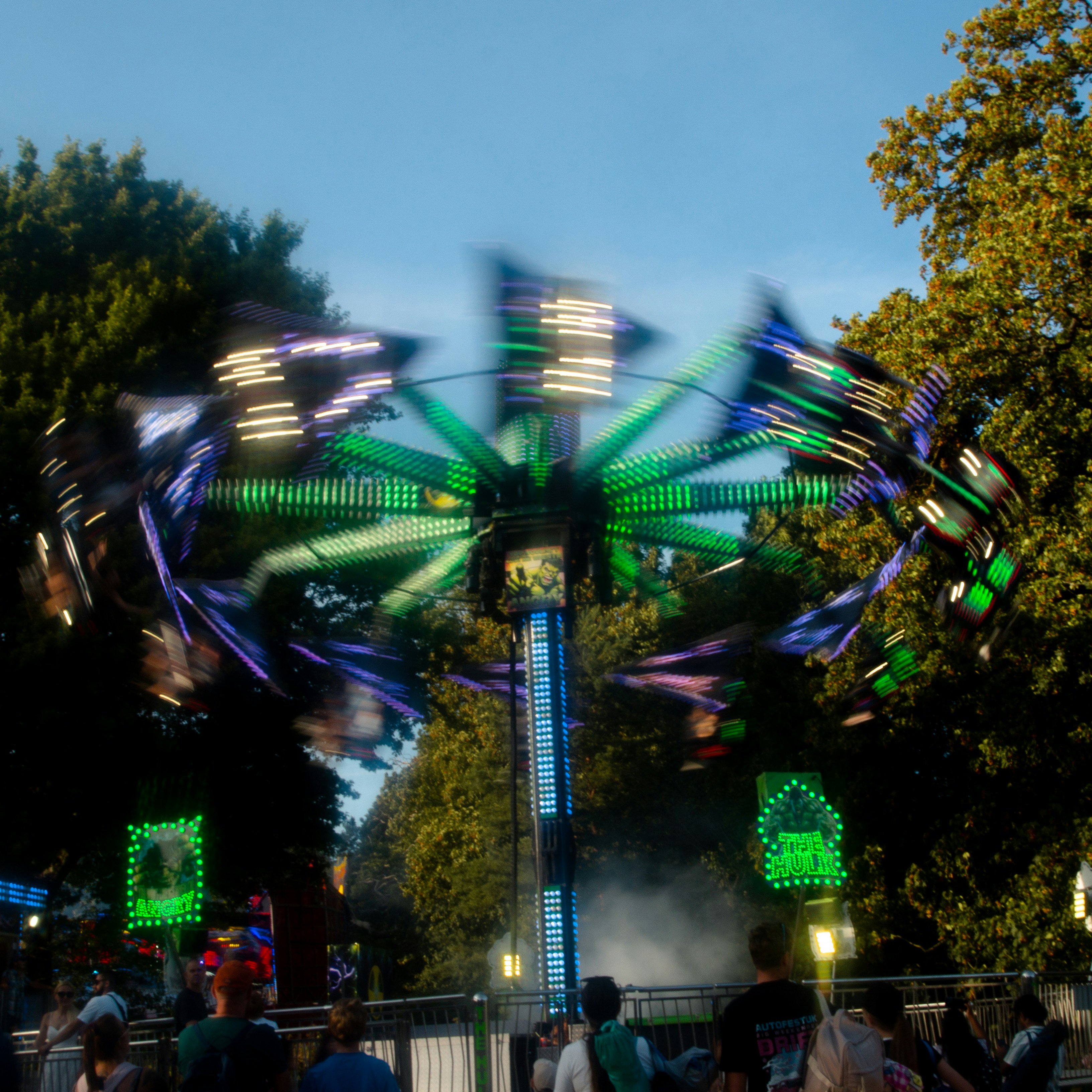 A blurred amusement ride spins under trees with bright green and purple LEDs, capturing motion and energy at a daytime fairground. This image emphasizes dynamic color trails and atmospheric lights.