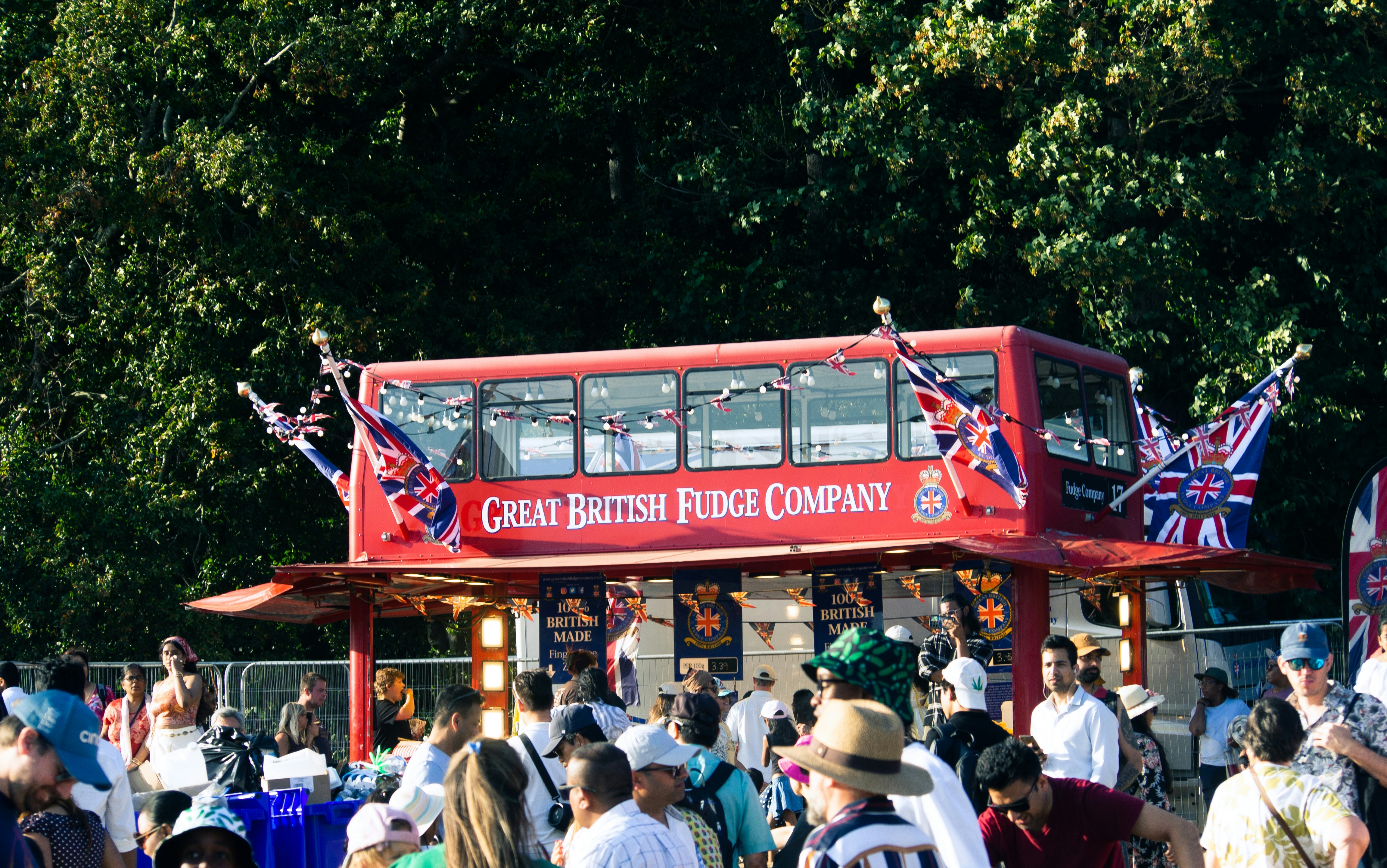 Bright red double-deck bus with 'Great British Fudge Company' signage amid a lively crowd at a festival; Union Jack banners and people in casual attire create a festive, sunlit scene.