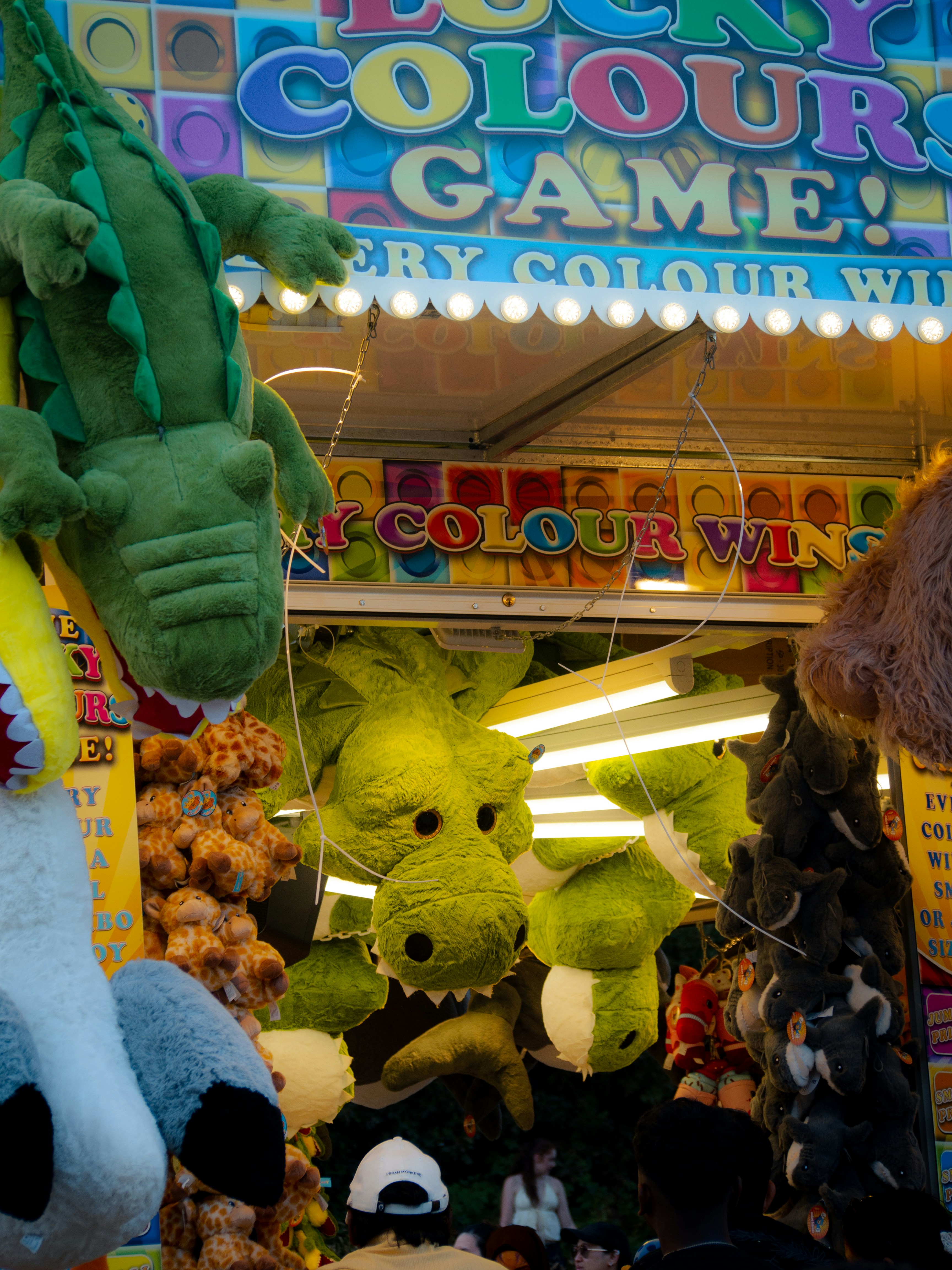 Vibrant carnival stall with oversized plush dragons and animals, colorful signage overhead, and a crowd below; a playful arcade backdrop for stuffed toy prizes.