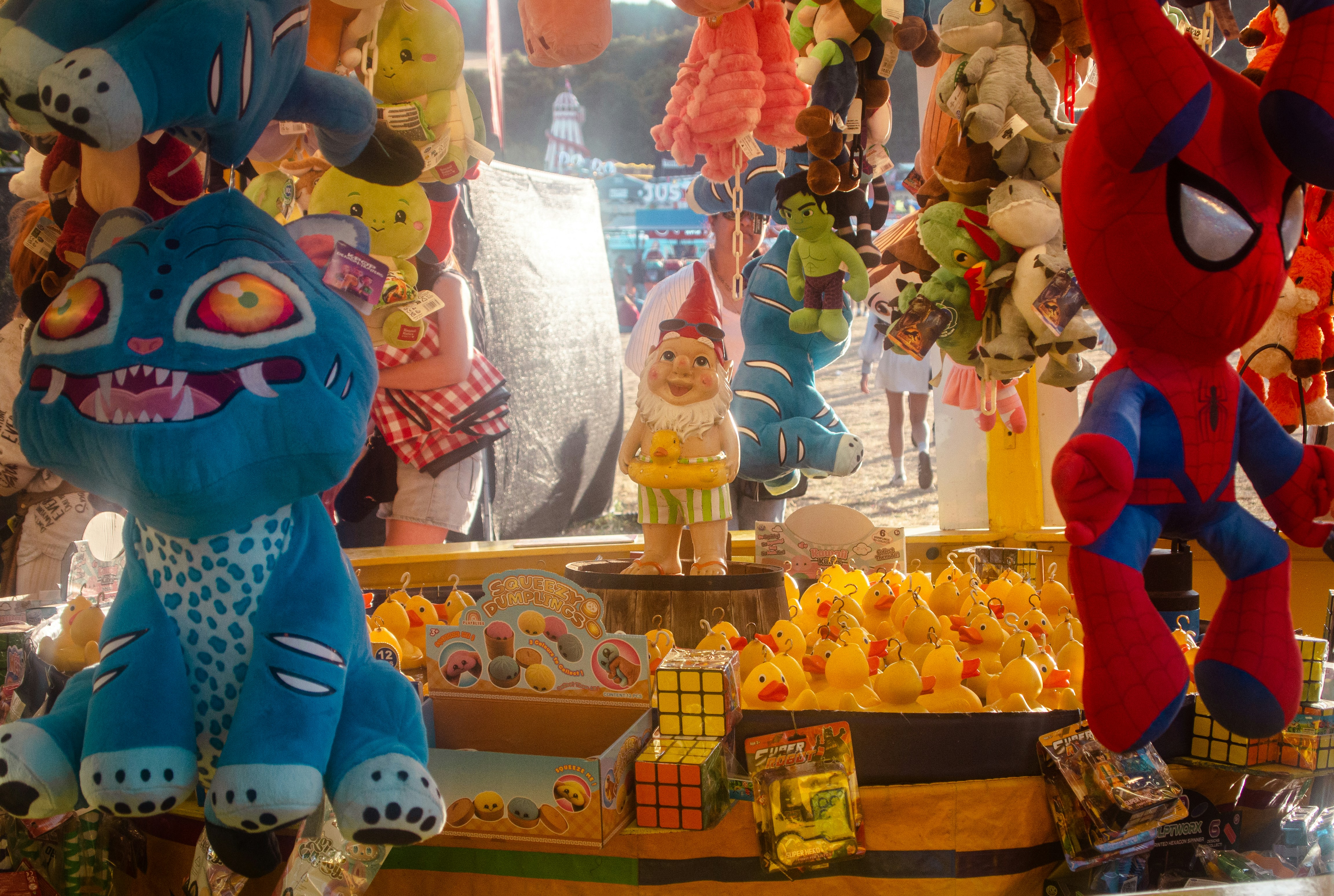 Colorful plush characters fill a carnival game stall, with stuffed animals and a Rubik’s cube display framing a central small figure; the scene conveys a busy prize booth at a fair.