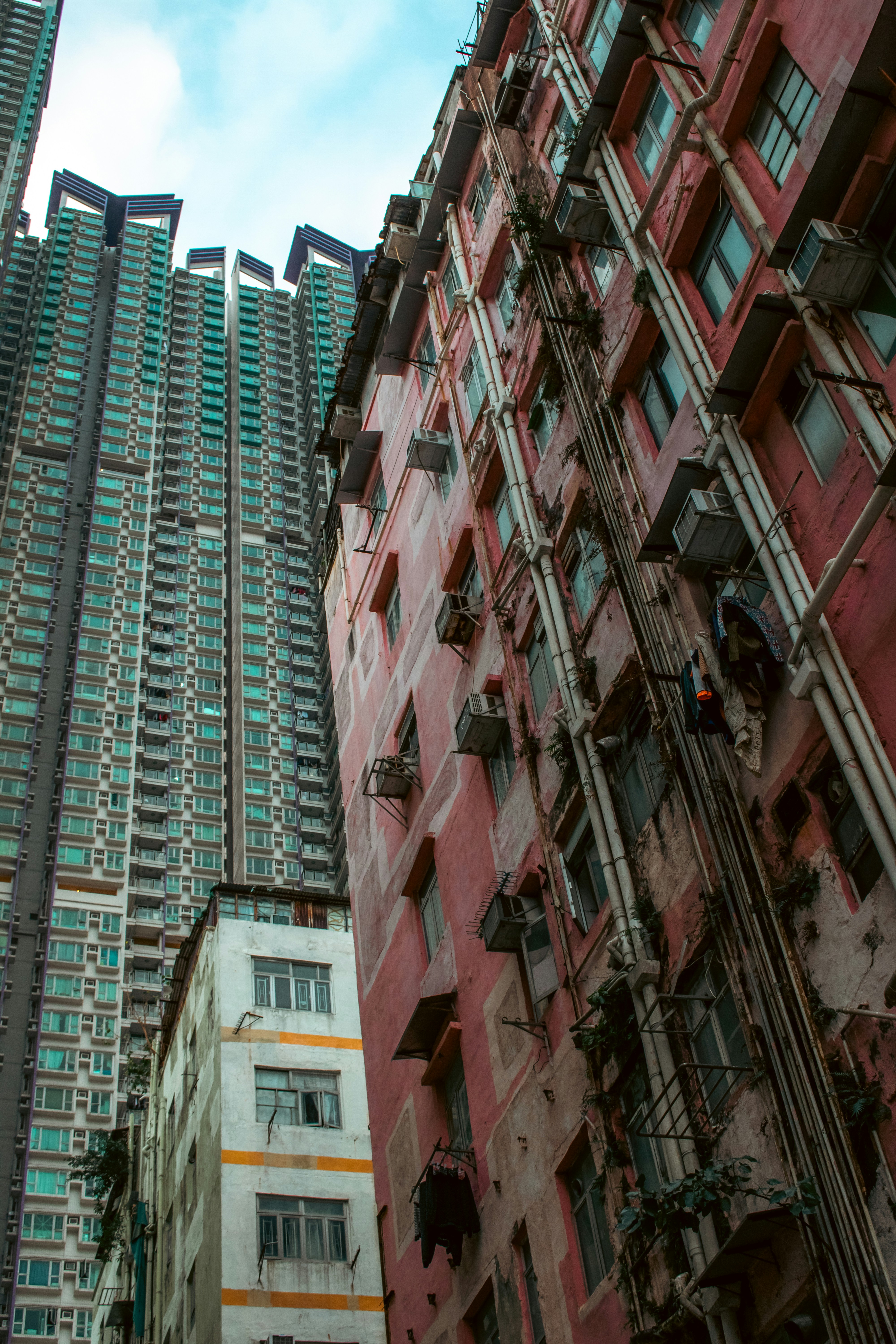 In Kai Tak, Hong Kong, aging low-rise buildings sit dwarfed in the shadow of new high-rises, framed from a narrow alley below. The contrast traces back to Kai Tak Airport. While it operated, flight paths capped building heights across the area. When the airport closed, those restrictions lifted, and towers shot up around the old low-rises that remain.