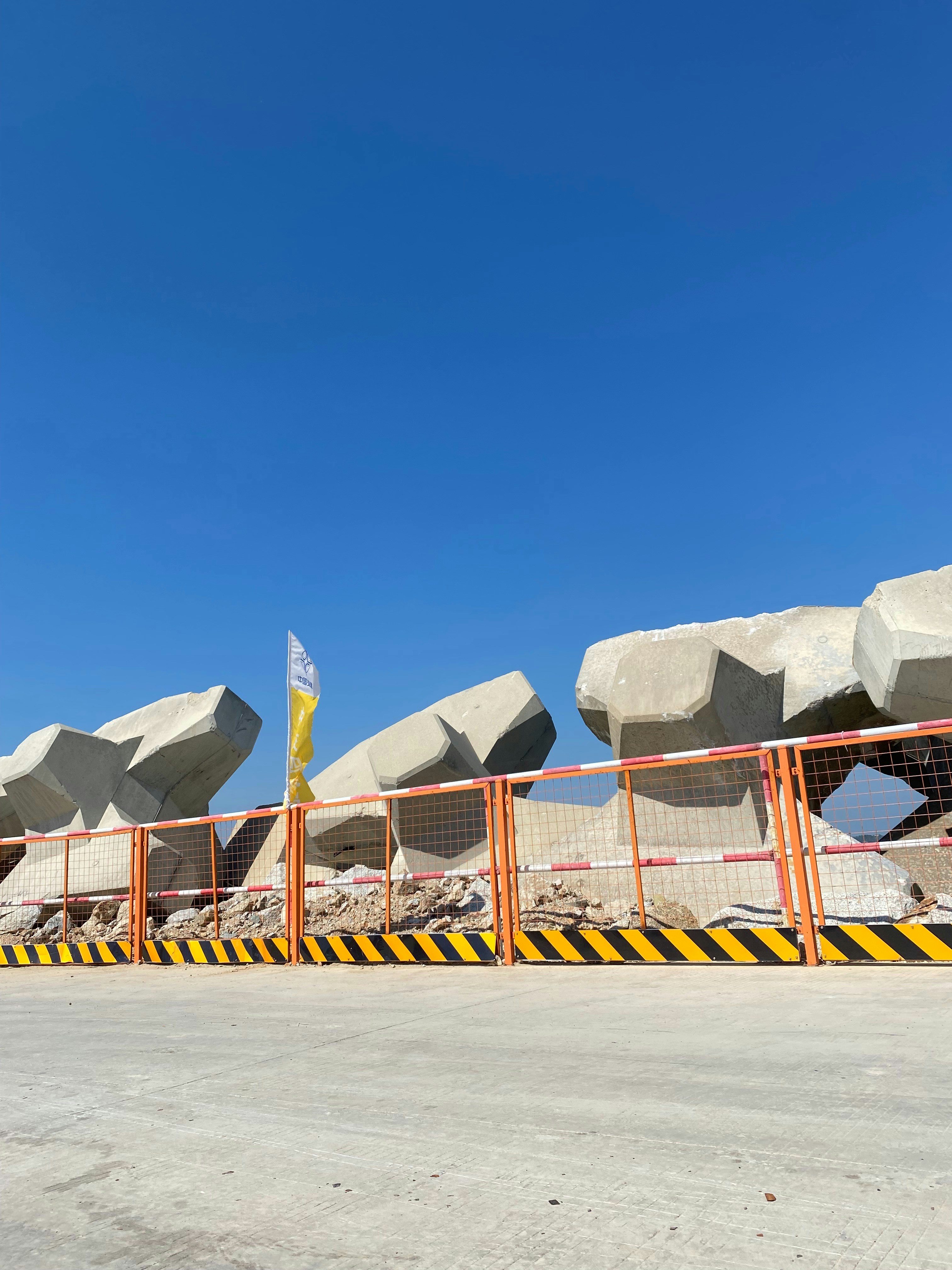 Concrete breakwater blocks behind orange safety fencing set on a sunny dock, with a clear blue sky above and construction elements in the foreground.
