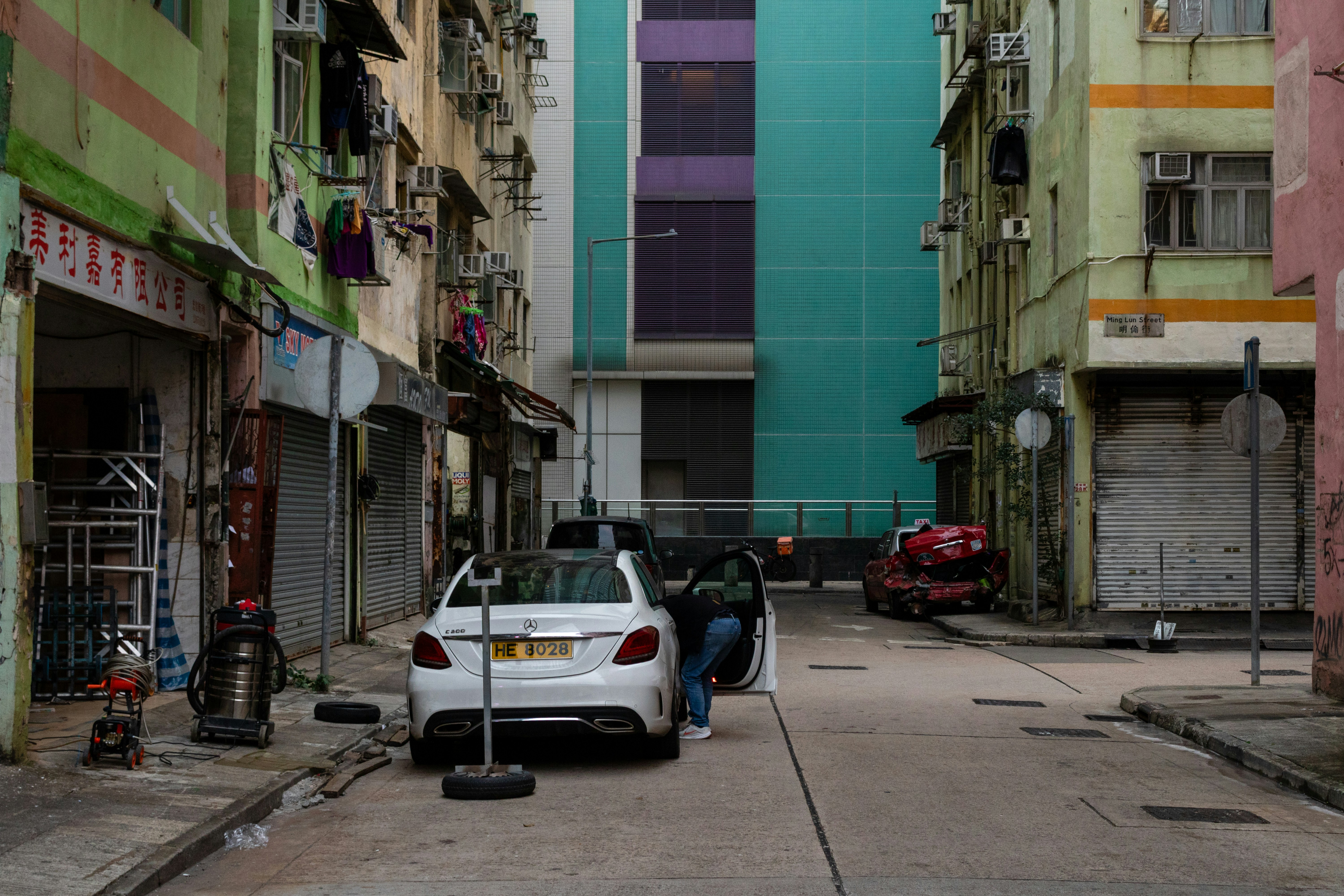 Kai Tak, one of Hong Kong’s older urban districts, showing aging low rise buildings, colorful façades, traditional shopfronts, exposed air conditioning units, and a modern turquoise building rising behind them. A white Mercedes is parked in the street beside automotive equipment, while a damaged red taxi sits further down the road.