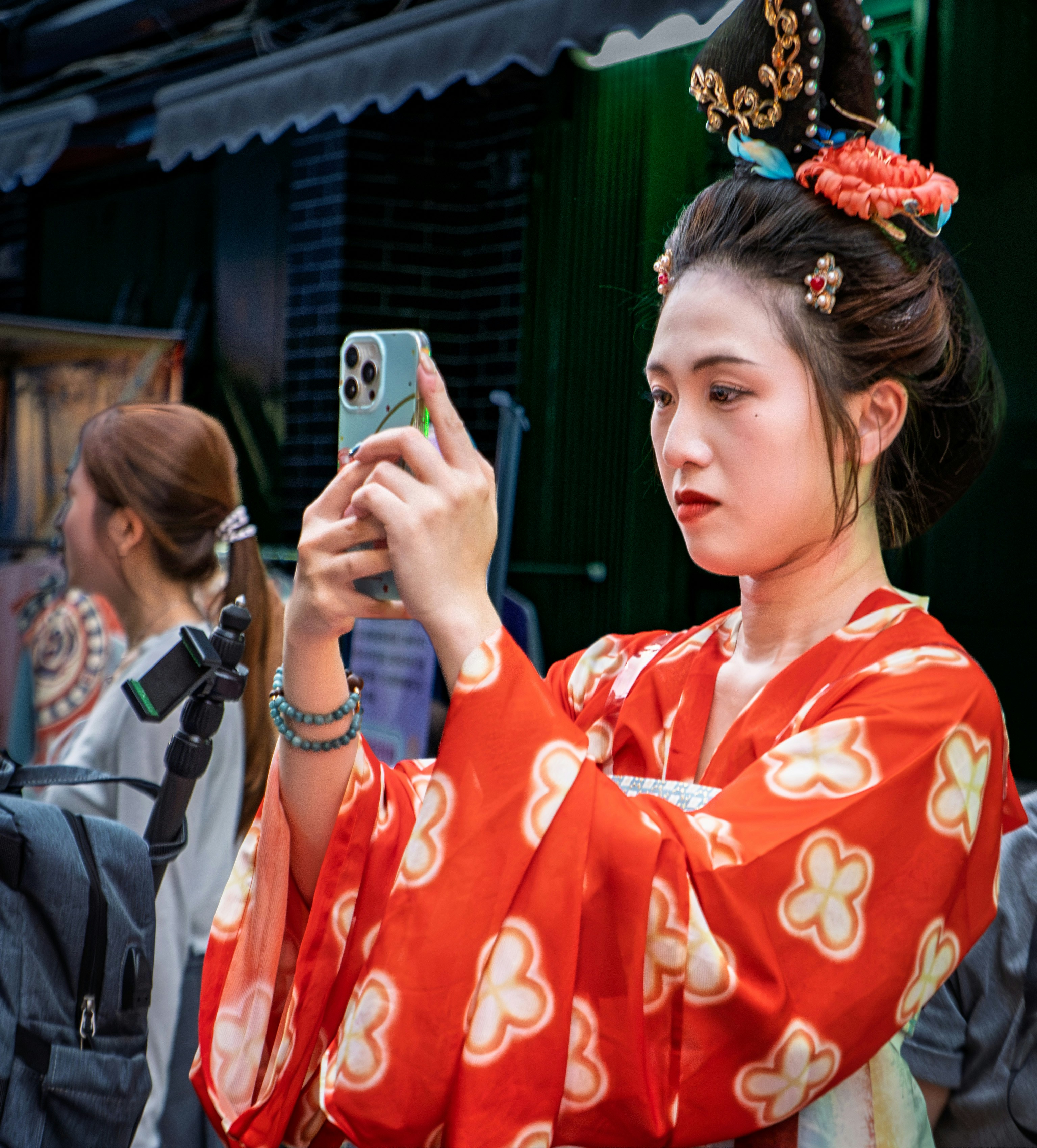 A woman in a vibrant red kimono stands on a busy street, focused on her smartphone as she records or photos documents; the scene includes passersby and urban elements in the background.