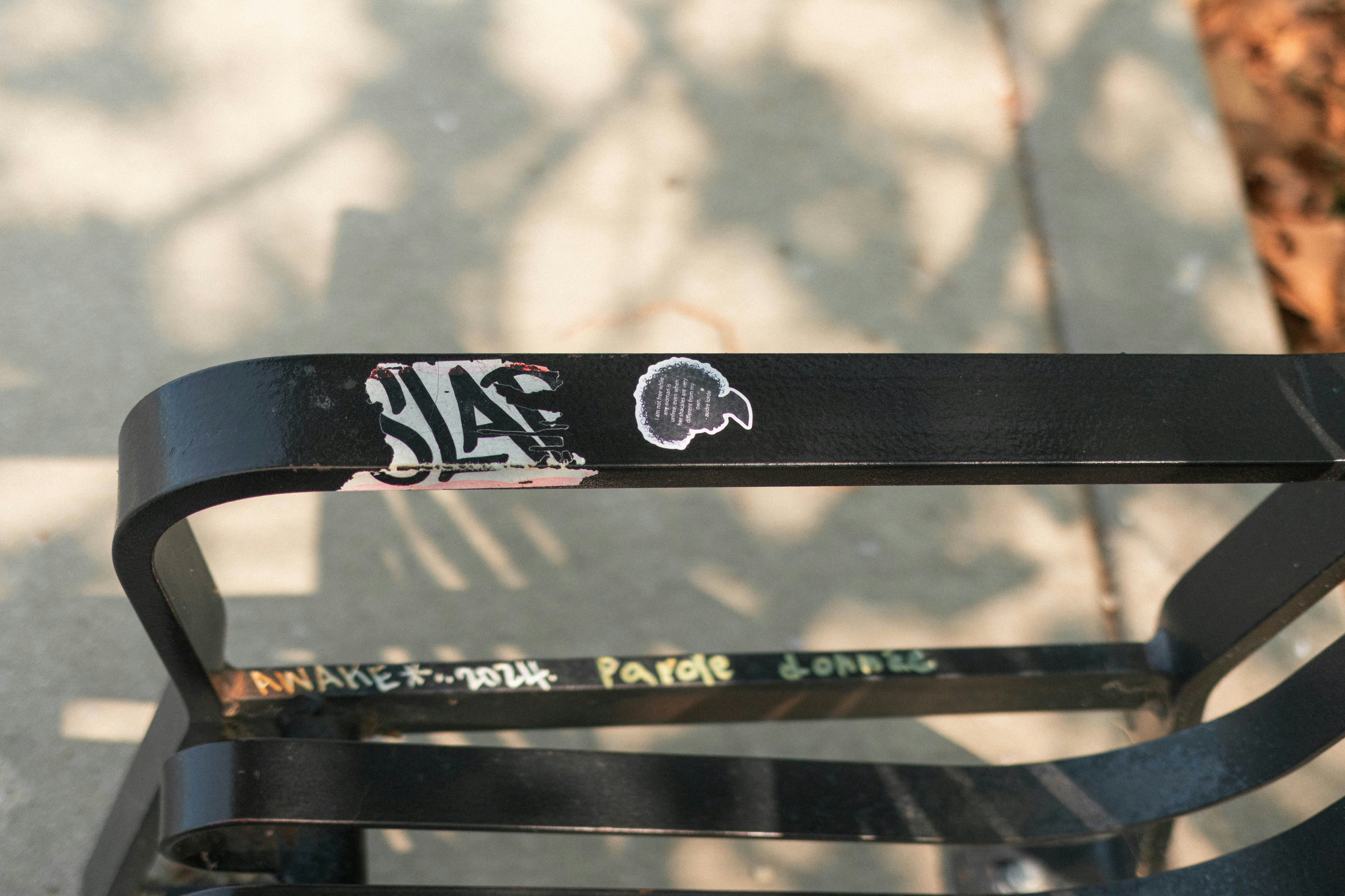 Close-up of a black metal park bench railing with stickers and scuffs; soft, dappled shadow pattern on concrete behind, suggesting an outdoor urban setting.