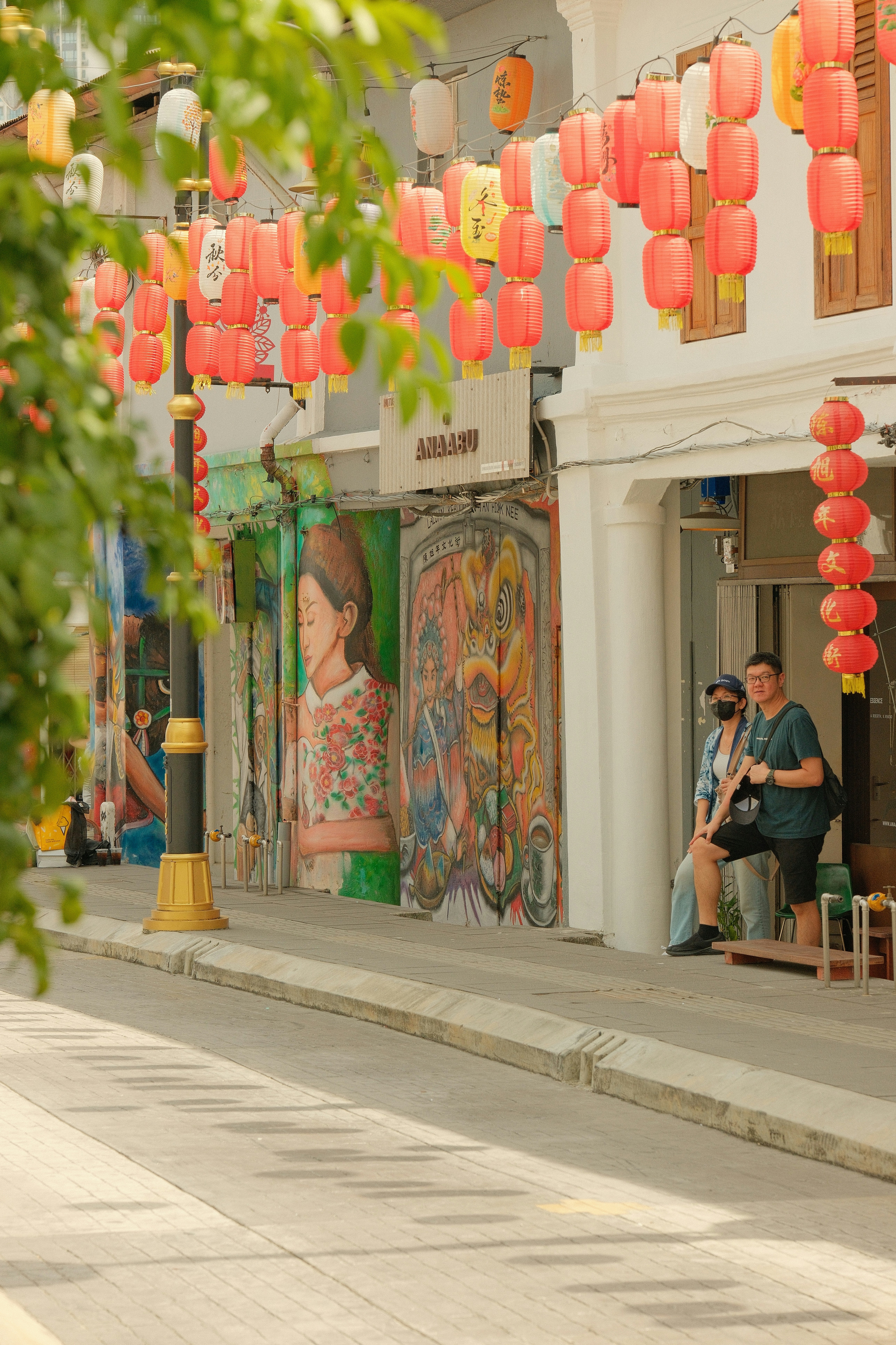 Quiet street scene with red lanterns overhead and a muraled storefront; two people sit beside the entrance, a painterly mural dominates the wall, and soft daylight bathes the scene.