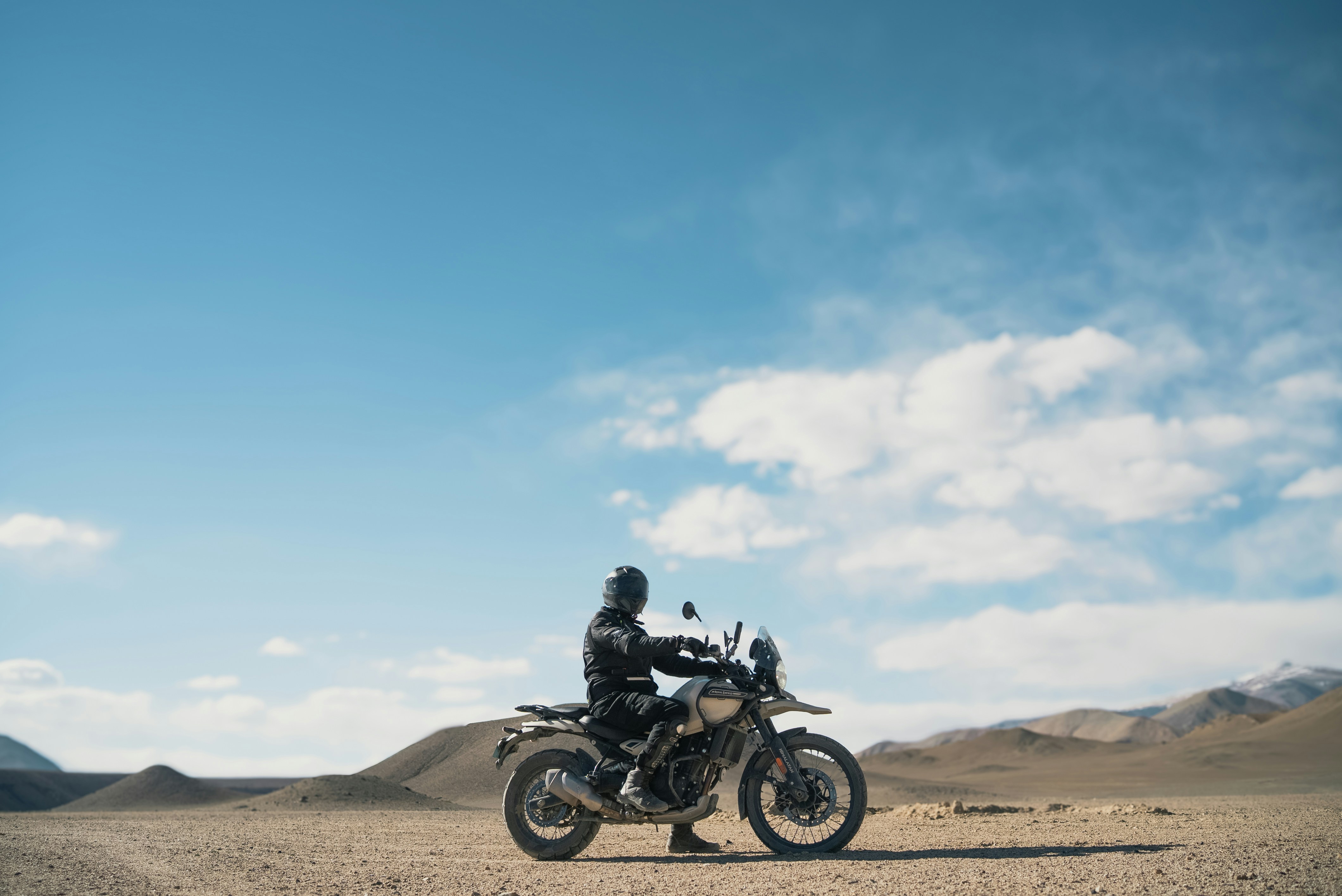 Rider on a dual-sport motorcycle traverses a barren desert with distant dunes under a bright sky. A titled composition emphasizes the lone figure and vehicle against expansive, arid terrain.