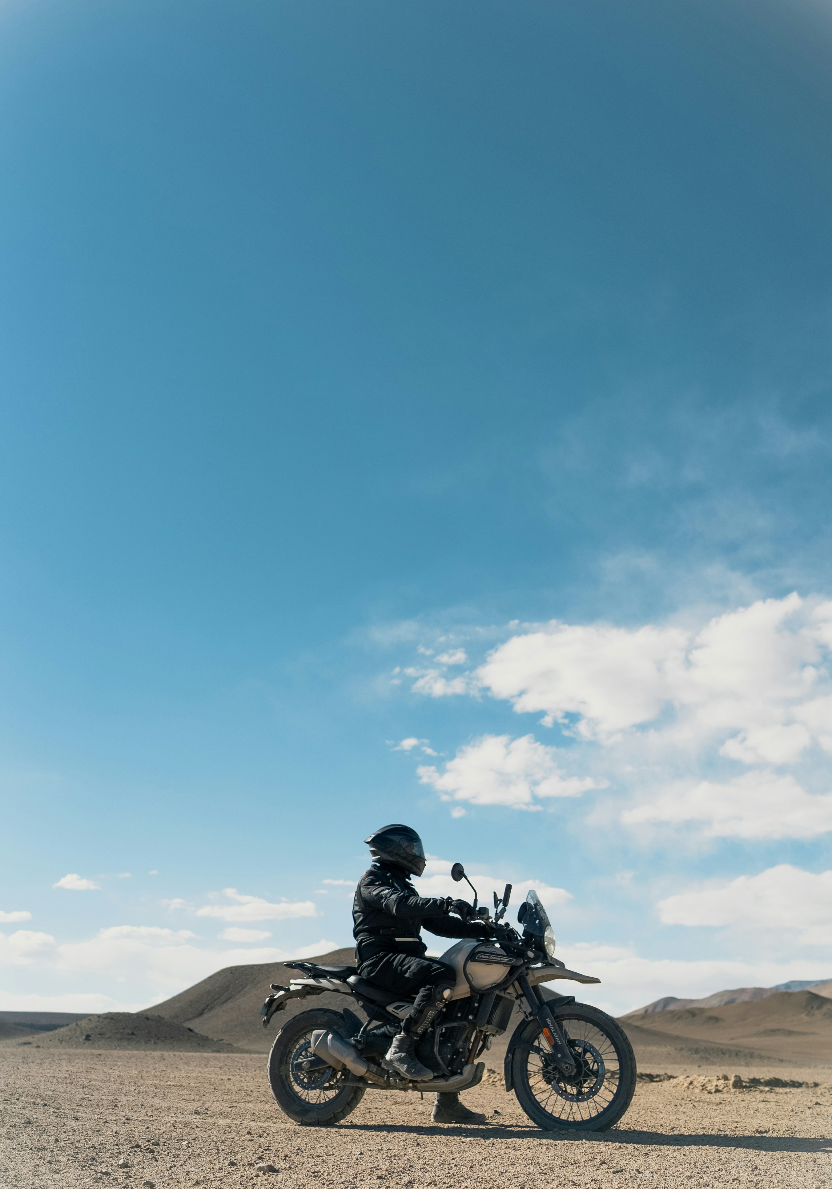 Motorcycle rider in full gear sits on a rugged desert bike beneath a vast blue sky with scattered clouds. The scene emphasizes solitude and open space in a stark, sunlit landscape.