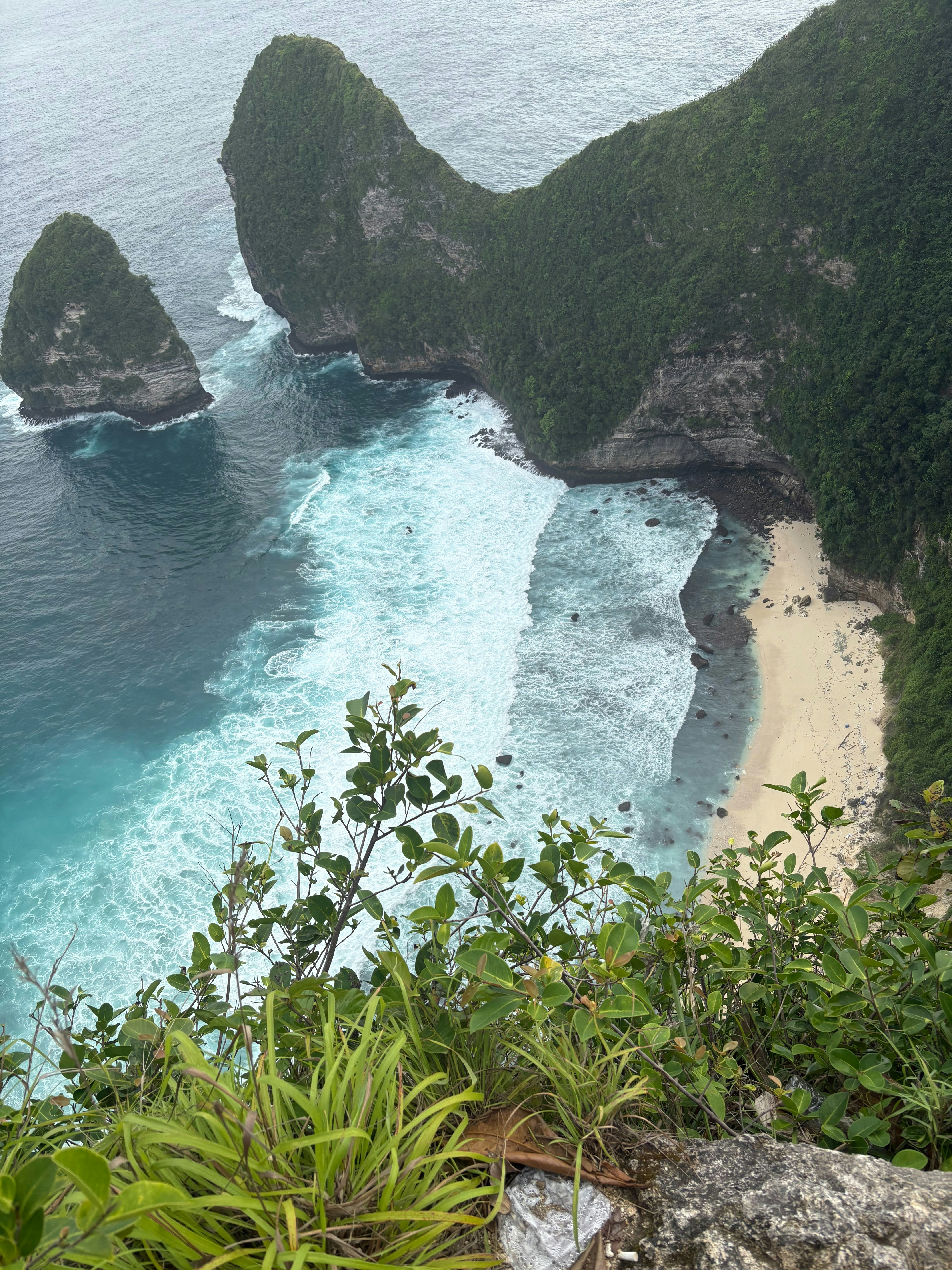 A rugged coastline viewed from a cliff, where turquoise surf meets a sandy cove flanked by green cliffs and coastal vegetation.
