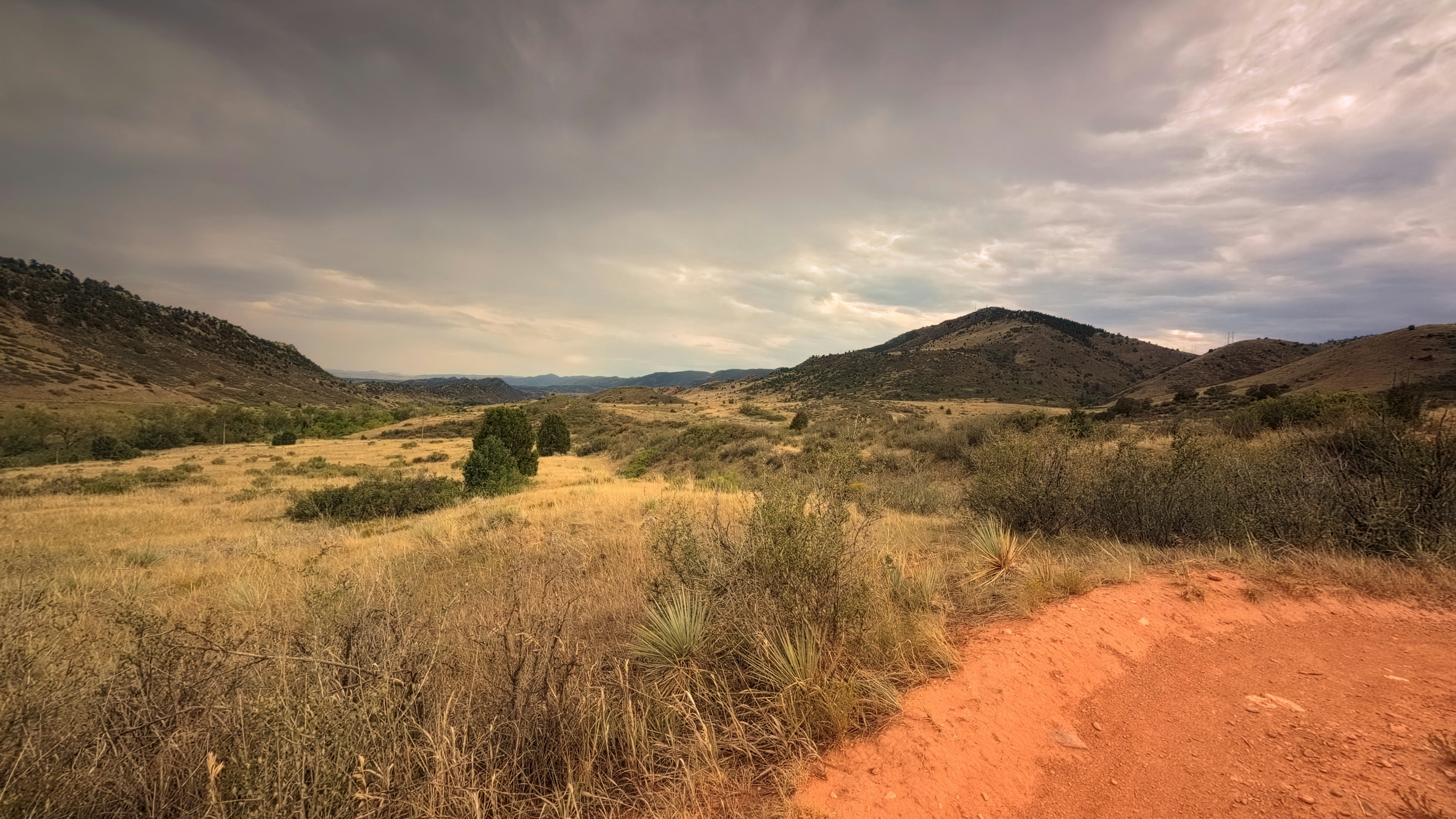 Dusty canyon trail winds toward distant hills with sparse shrubs under a moody sky.