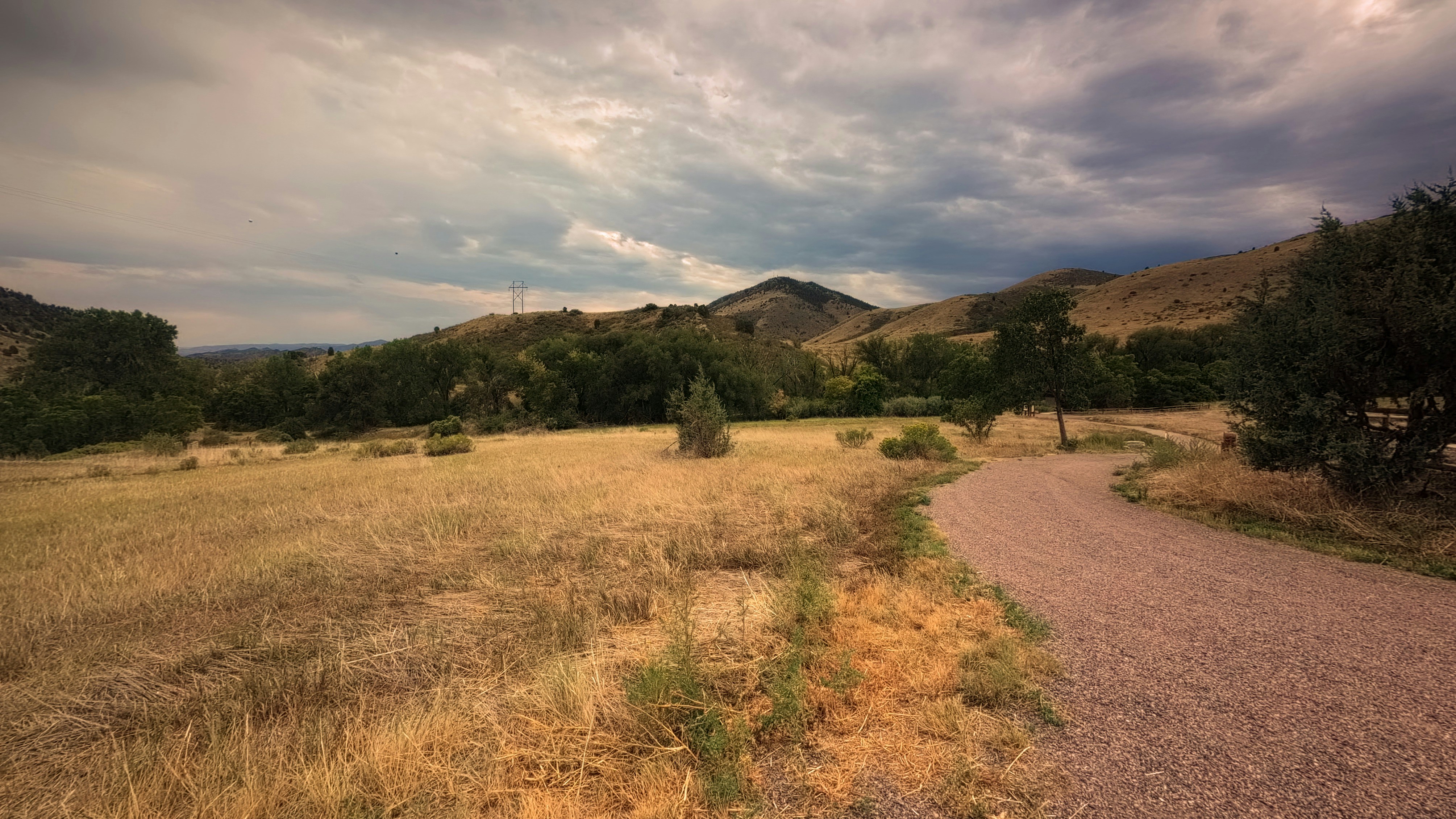 Dusty gravel path winds through a sunlit, arid landscape with scattered trees and rolling hills under a dramatic sky.