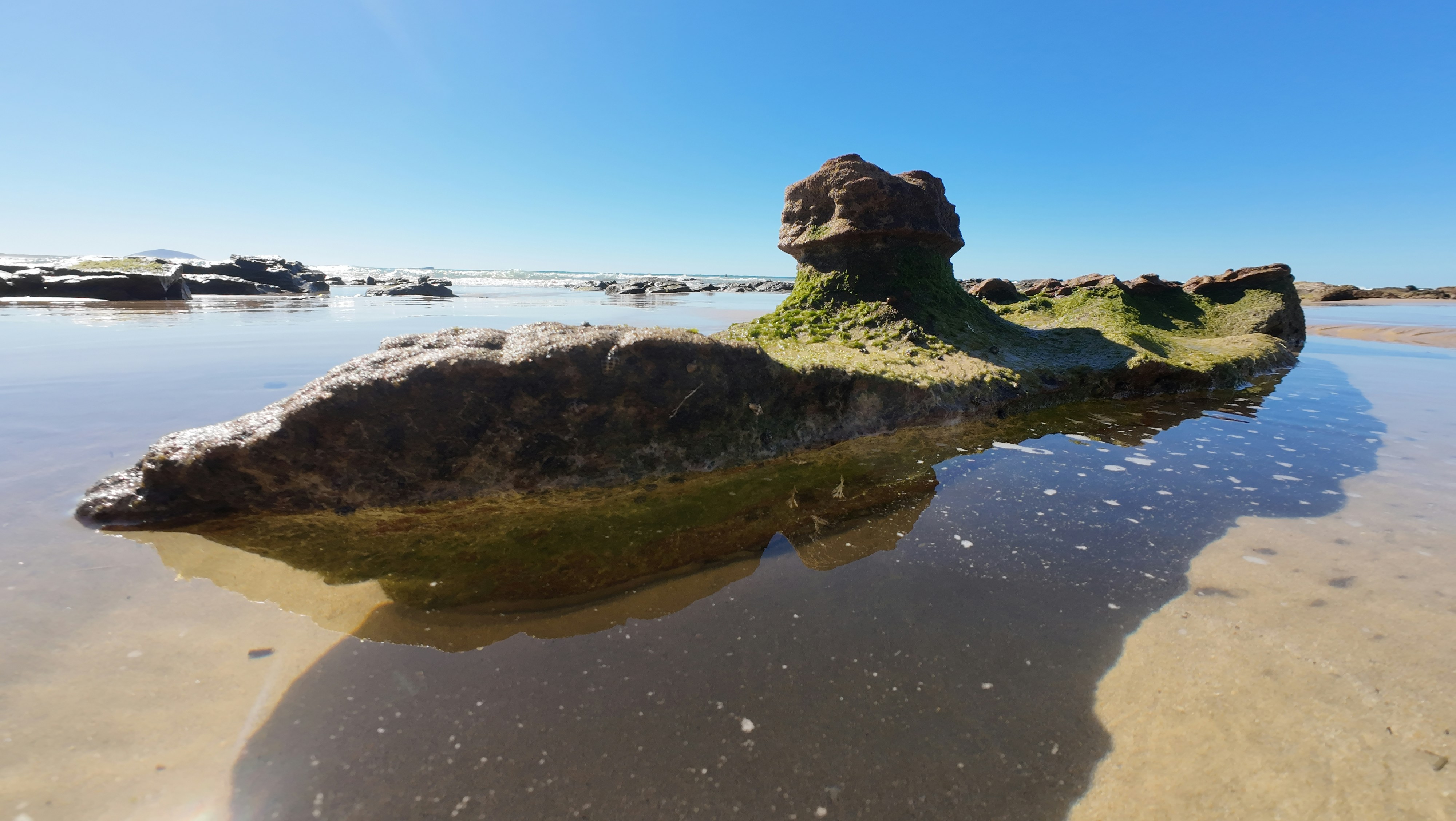 Rock formations rise above shallow tidal pools along a clear shoreline under a blue sky. A mossy stack anchors the scene with reflections and gentle water textures.