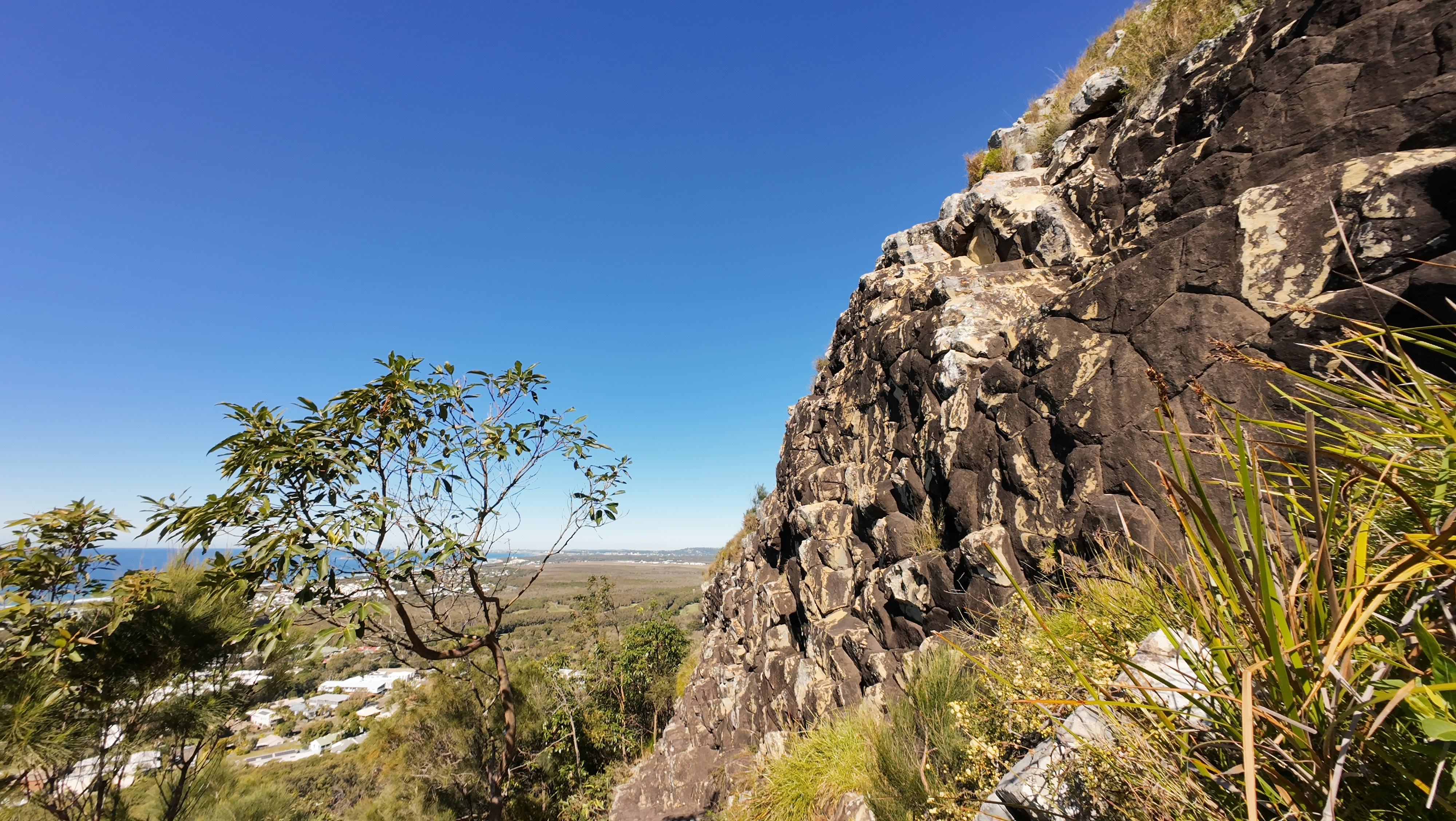 Cliff face rises on the right with sparse vegetation and a distant coastline under a bright sky.