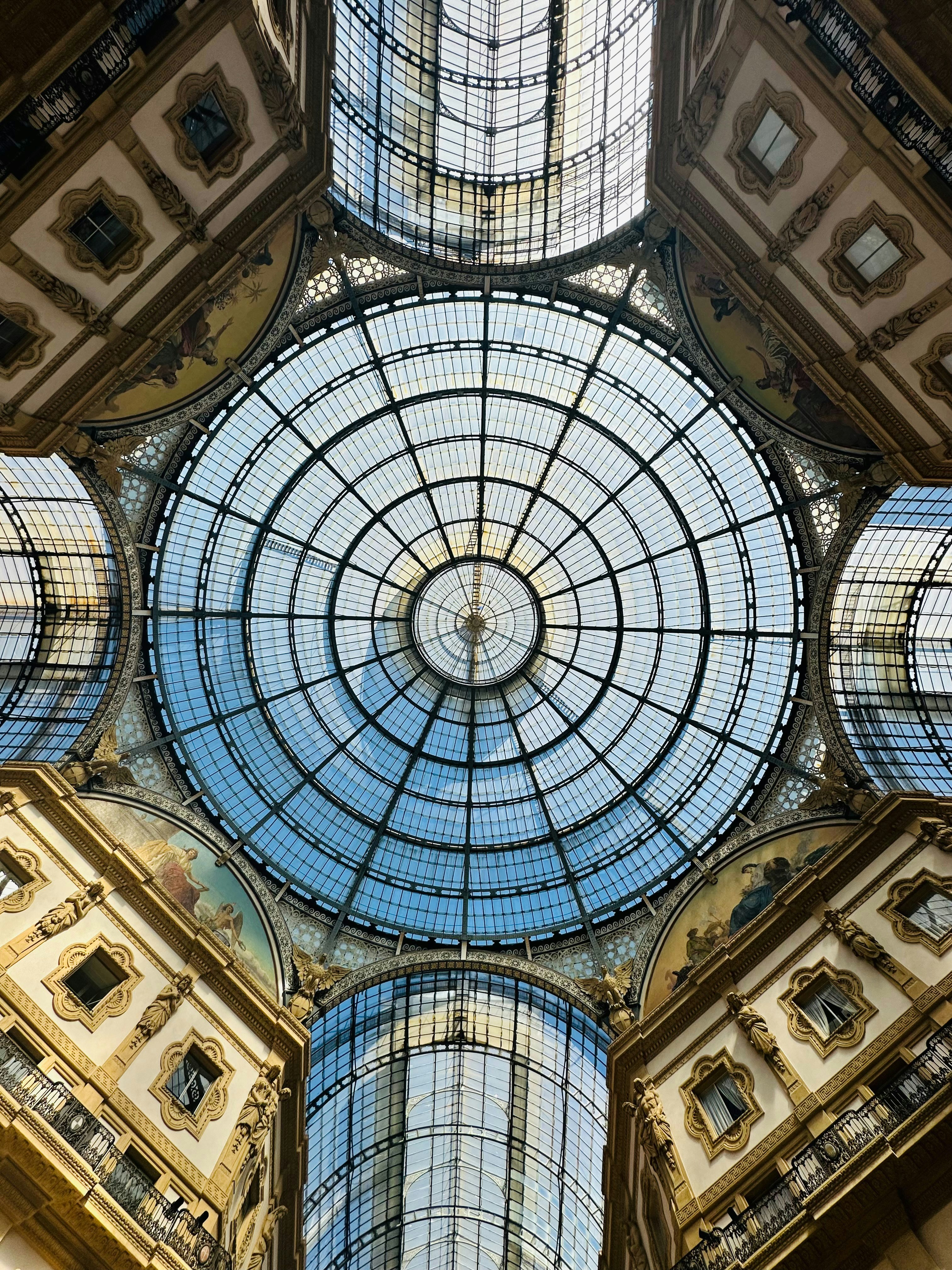 Intricate glass dome framed by classical architecture rises overhead, showcasing a radial steel-and-glass pattern with blue sky peeking through. A dramatic atrium scene of architectural symmetry.
