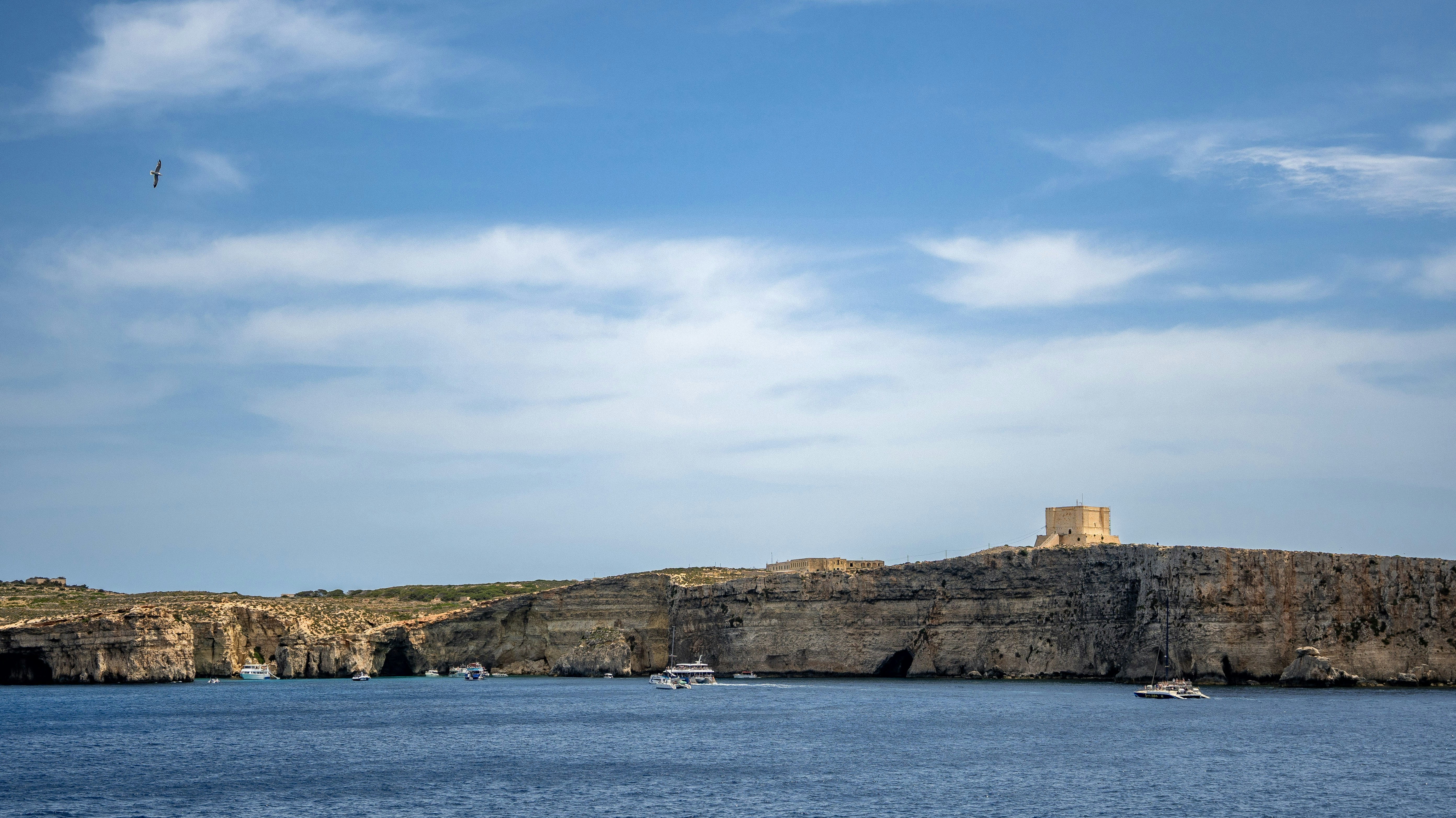 Roaring sea meets rugged cliff coast beneath a bright sky; a distant fortress sits atop the limestone escarpment as small boats glide by.