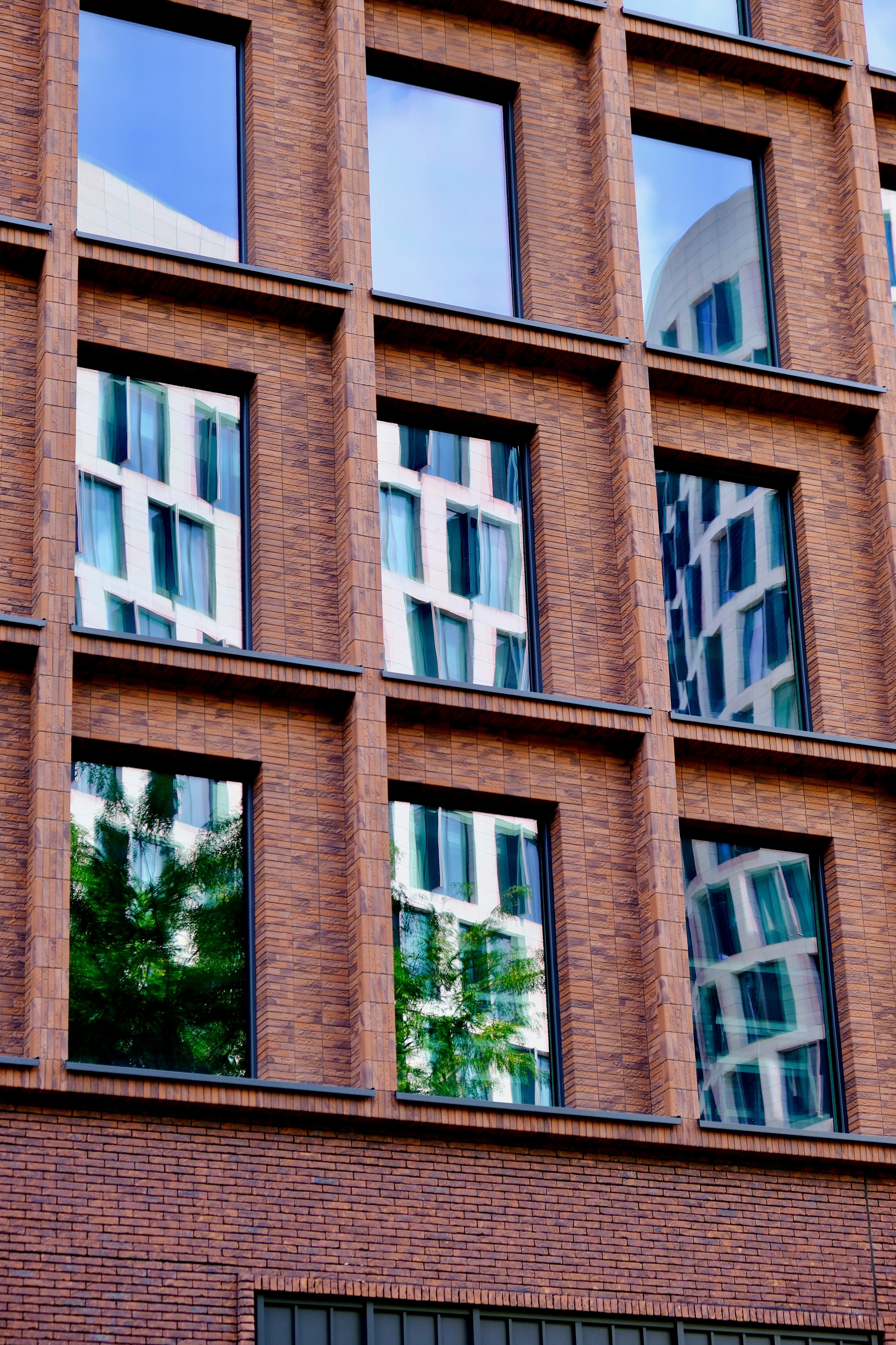 Brick-faced building with a grid of rectangular windows reflecting blue sky and surrounding structures; the pattern emphasizes geometry and repetition. The image showcases architectural detail and reflective glass surfaces.
