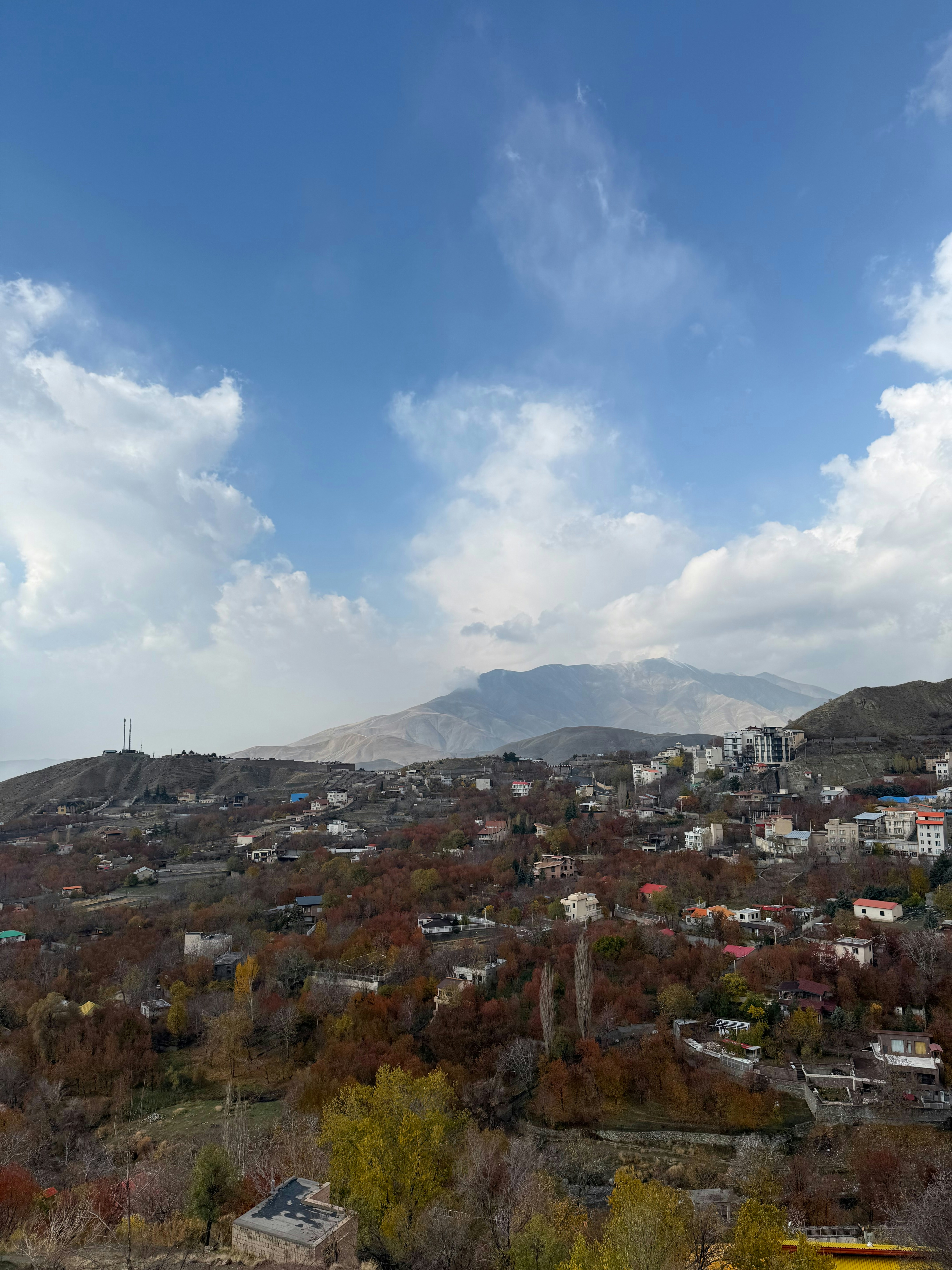 Autumnal townscape with a valley of houses and trees, backed by distant mountains and a bright blue sky with scattered clouds.