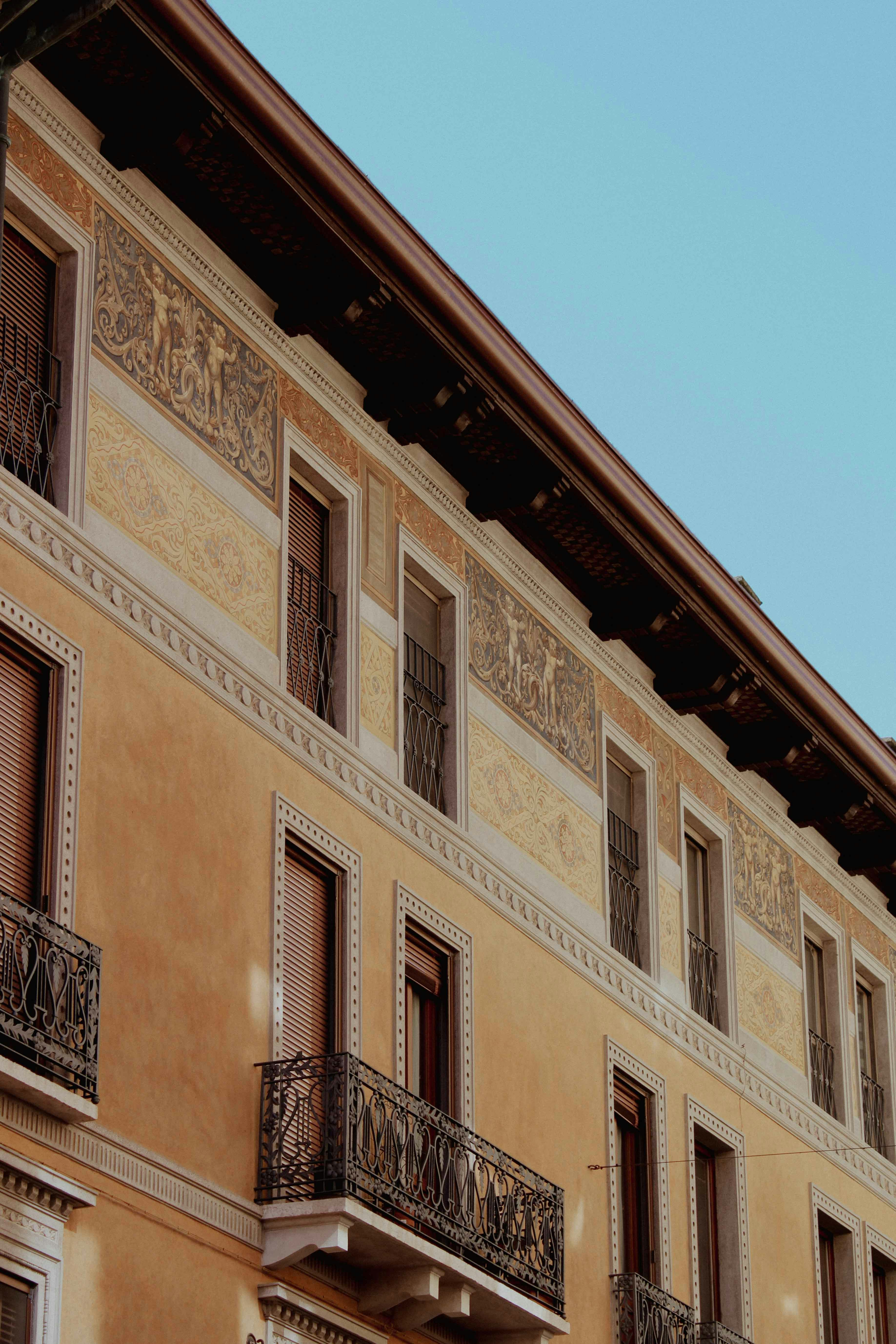 A warm, sunlit building facade with ornate stucco details and decorative iron balconies; the shot focuses on architectural features and patterning against a clear sky. This is a photograph of a architectural exterior, capturing texture and color.