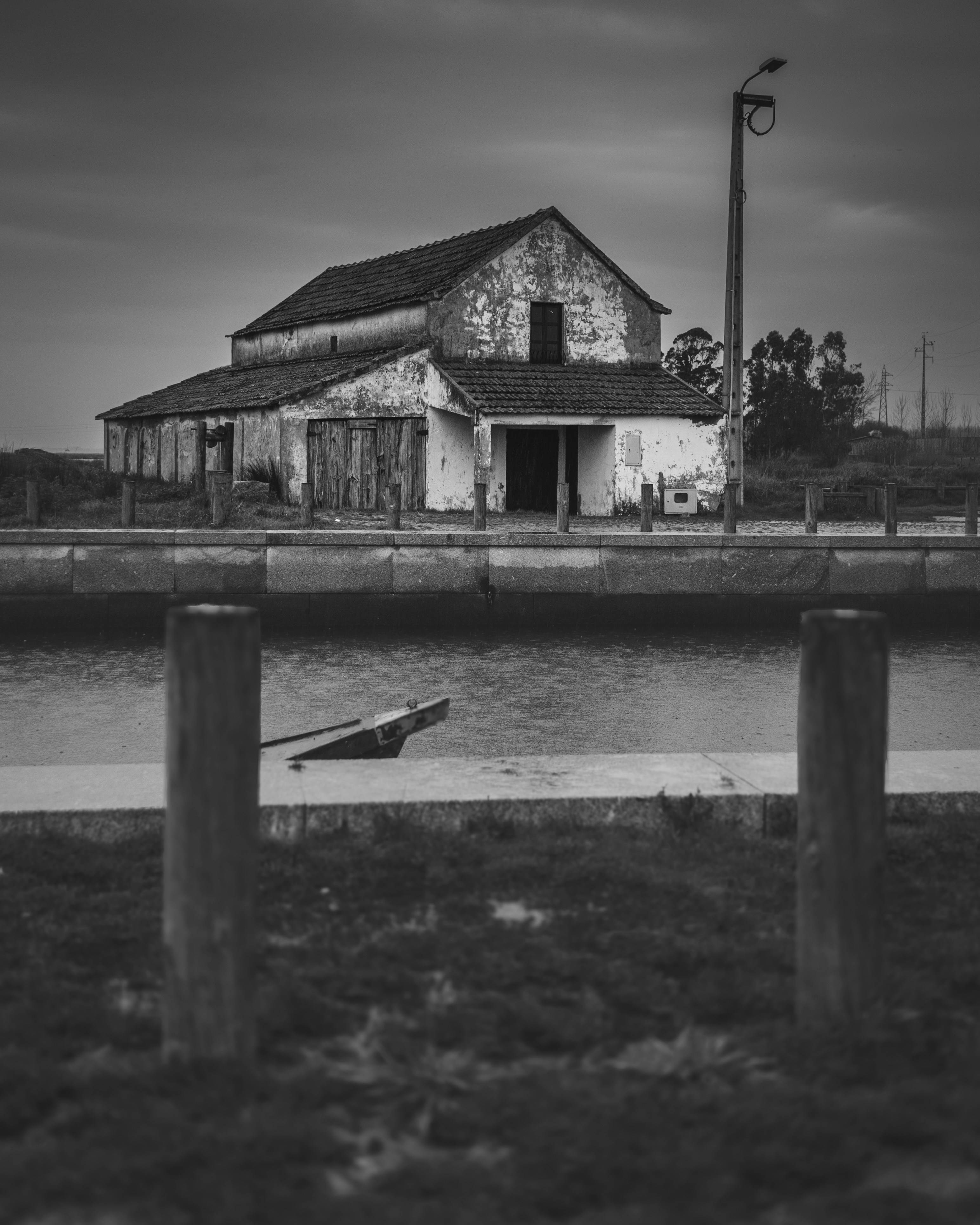 Weathered boathouse along a canal with a lone buoyant boat in foreground and posts lining the bank. Monochrome scene highlighting textures and decay.
