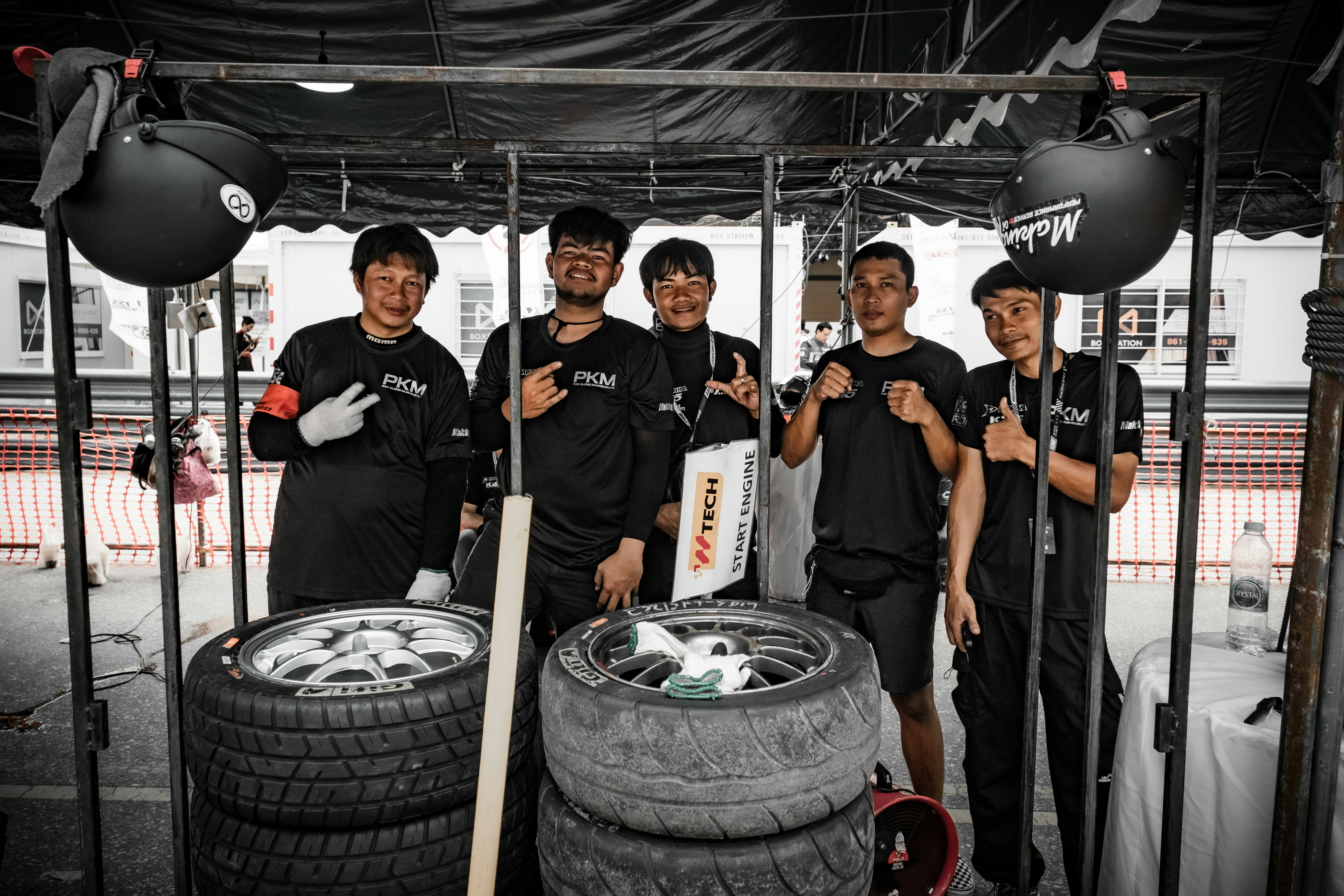An authentic, behind-the-scenes portrait of five motorsport crew members posing behind a metal equipment rack inside their pit tent at the Bangsaen Grand Prix. Wearing matching black PKM team shirts, the crew members give peace signs, thumbs-up, and hand gestures, surrounded by stacks of racing tires, safety helmets hanging from the steel frame, and technical pit gear. Captured during the Thailand Super Series event, this shot highlights paddock culture, mechanics, teamwork, and race weekend preparation.
