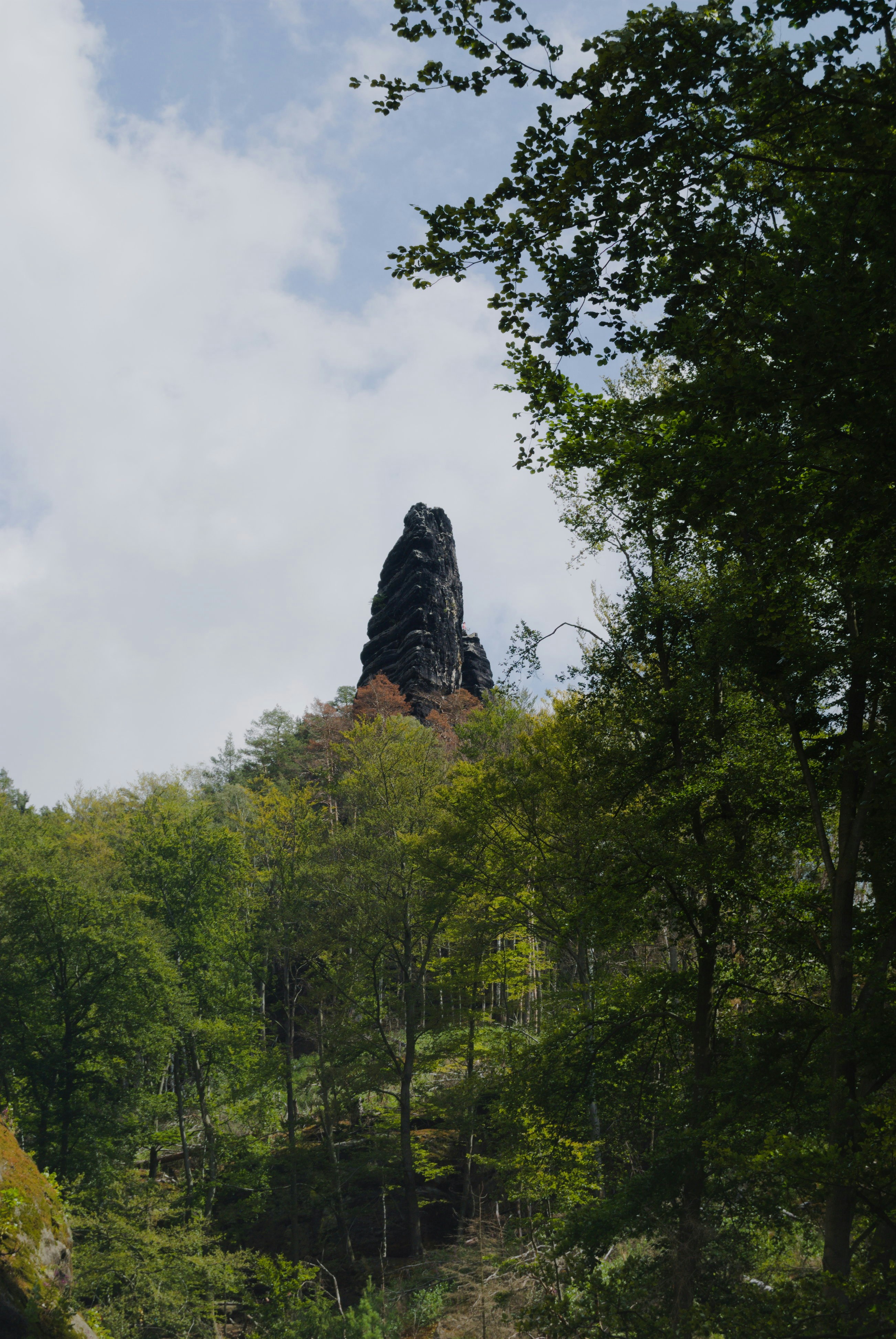 A jagged rock spire rises above a verdant hillside framed by trees and a bright sky.