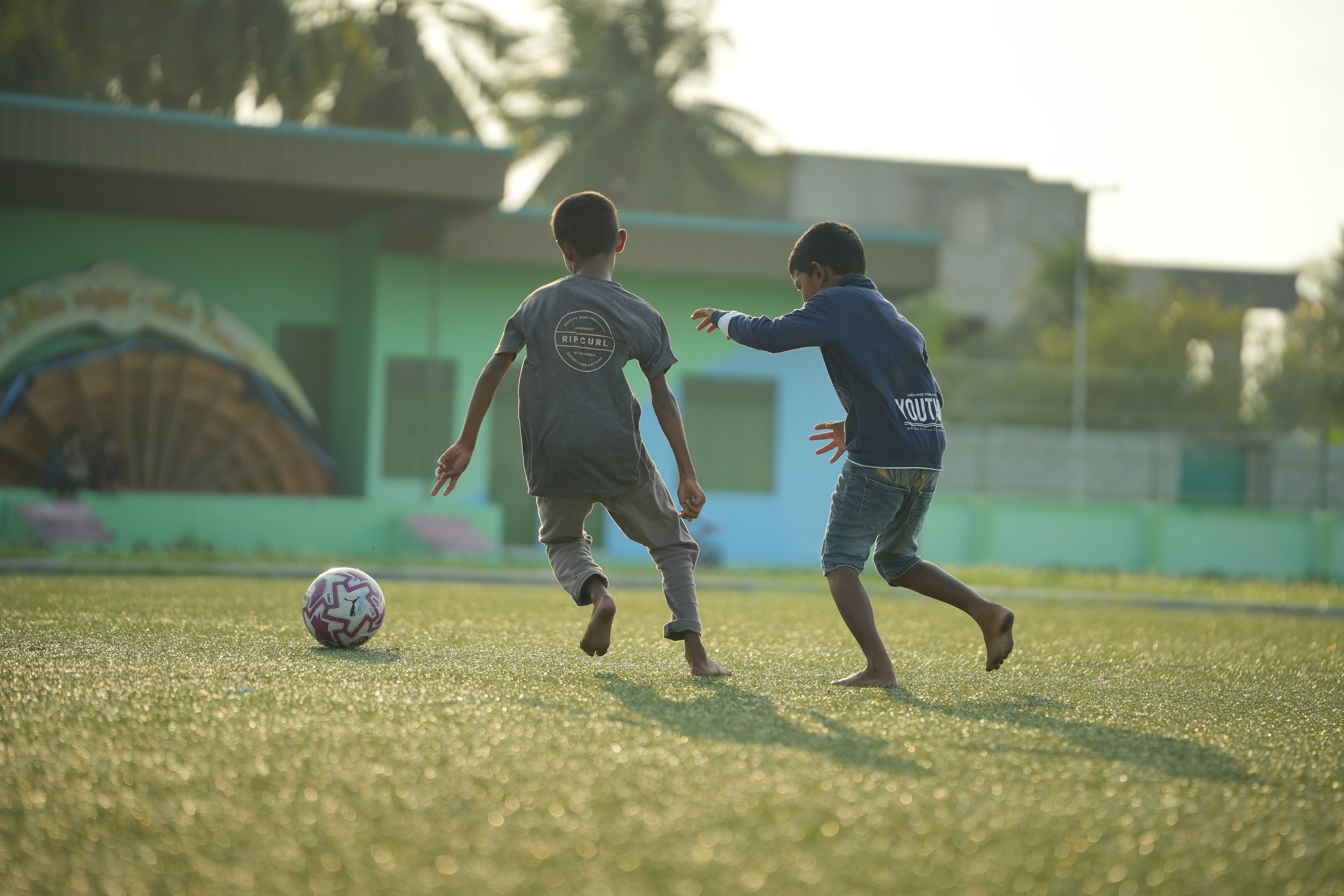 Two kids chase a soccer ball on a sunlit field at dusk