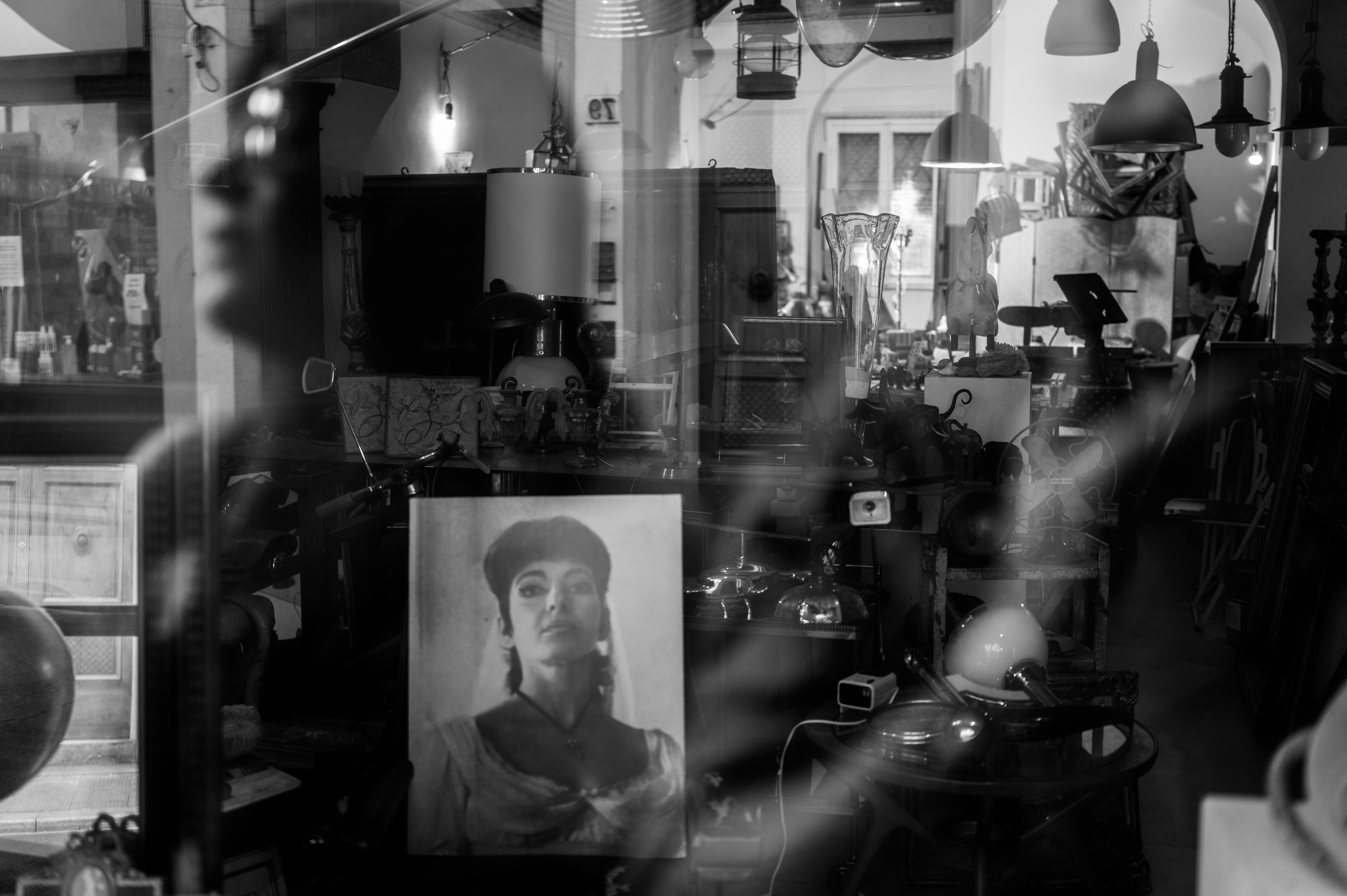 A grayscale scene captured through a shop window reveals a portrait placed amid an array of objects and shelves, creating a layered still-life in a candid interior setting.