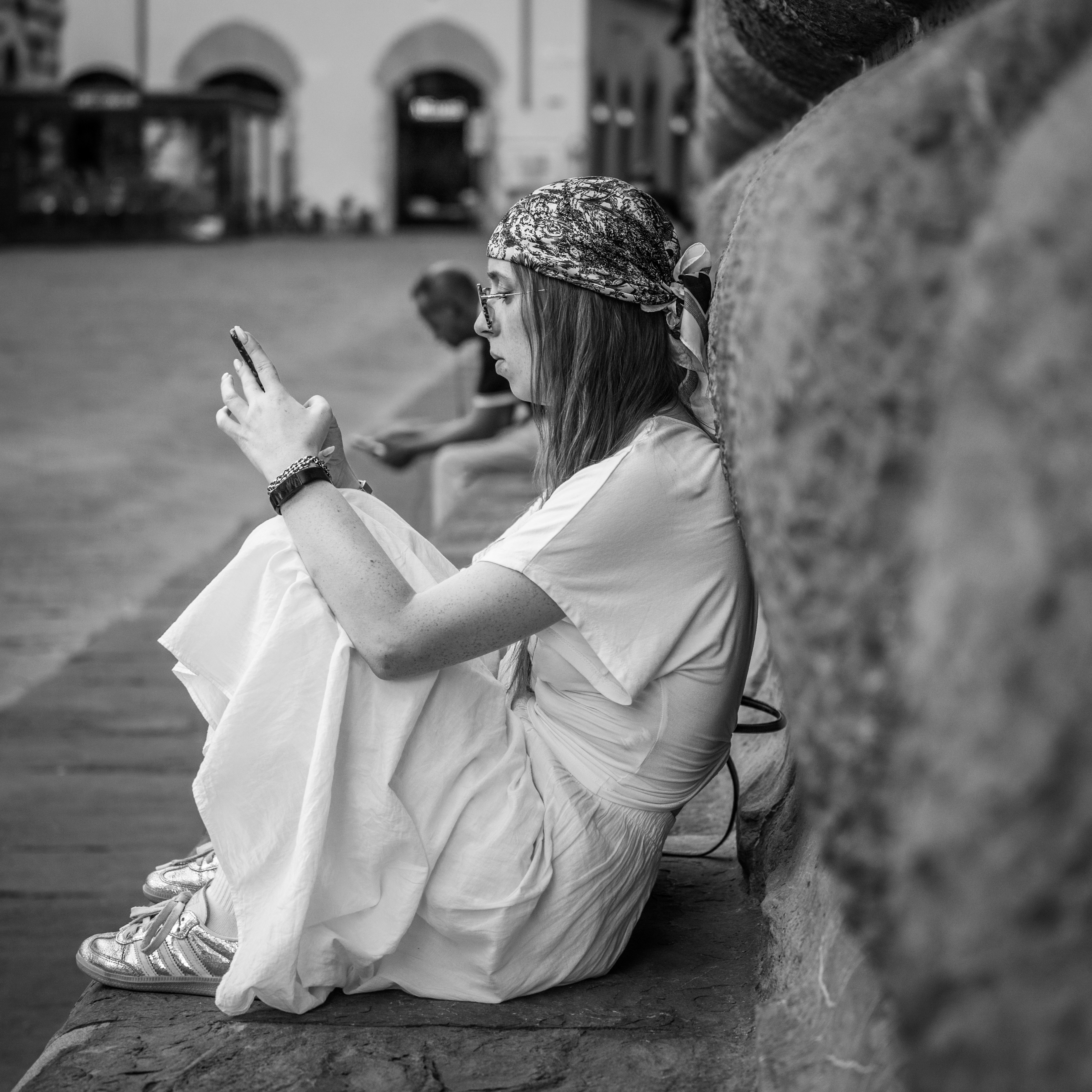 A woman sits on a stone ledge, absorbed in her smartphone, with a blurred background suggesting a public square; a monochrome portrait emphasizing texture and light.