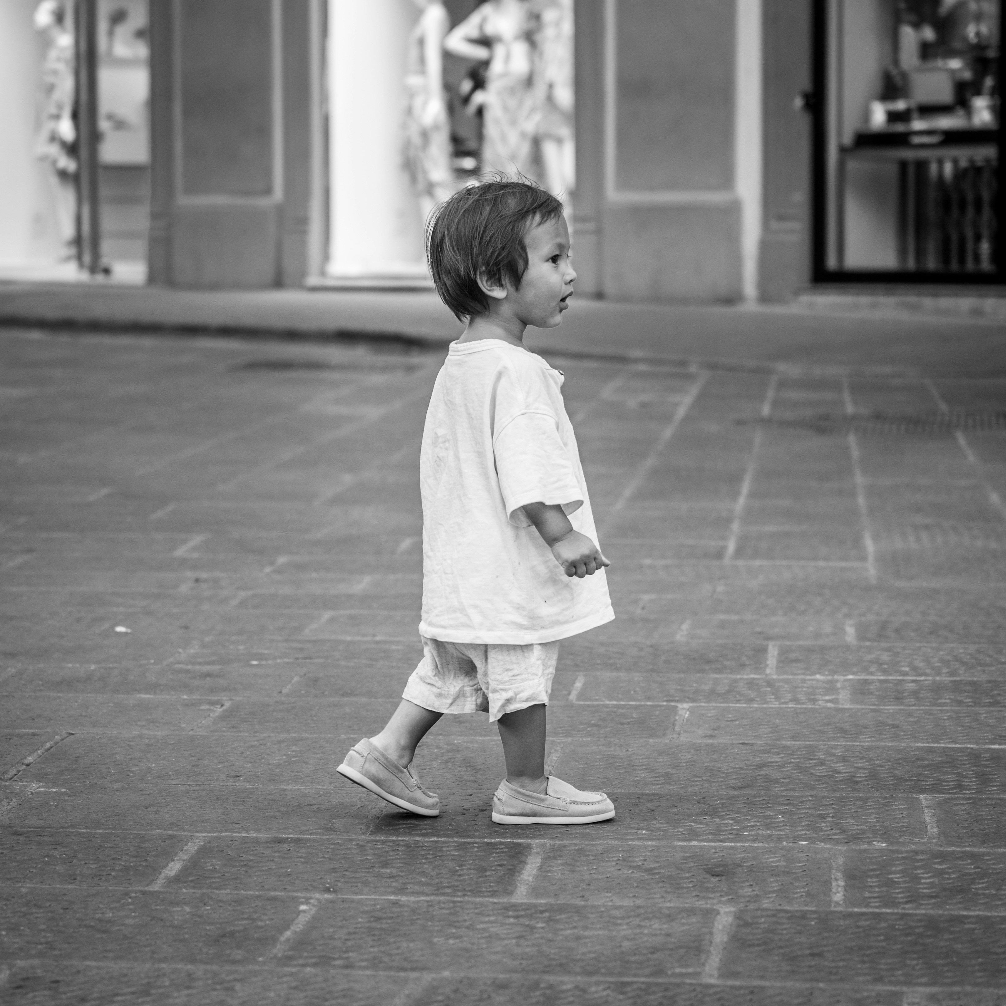 A grayscale street scene showing a small child walking along a stone-paved sidewalk, focused on the profile and casual attire as buildings frame the background.