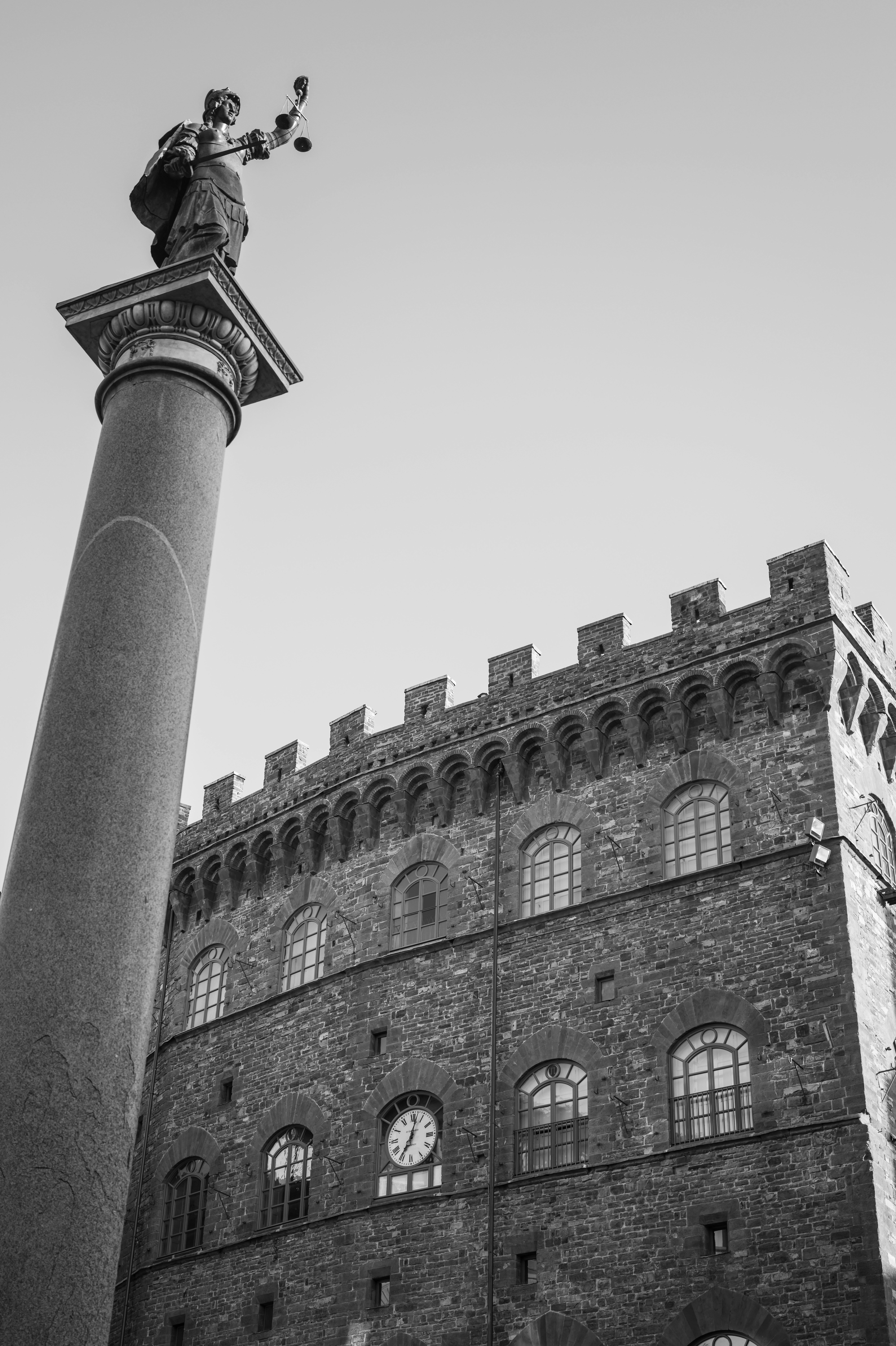 Tall column with statue atop and a stone building featuring a clock, captured in black and white to emphasize architecture and texture.