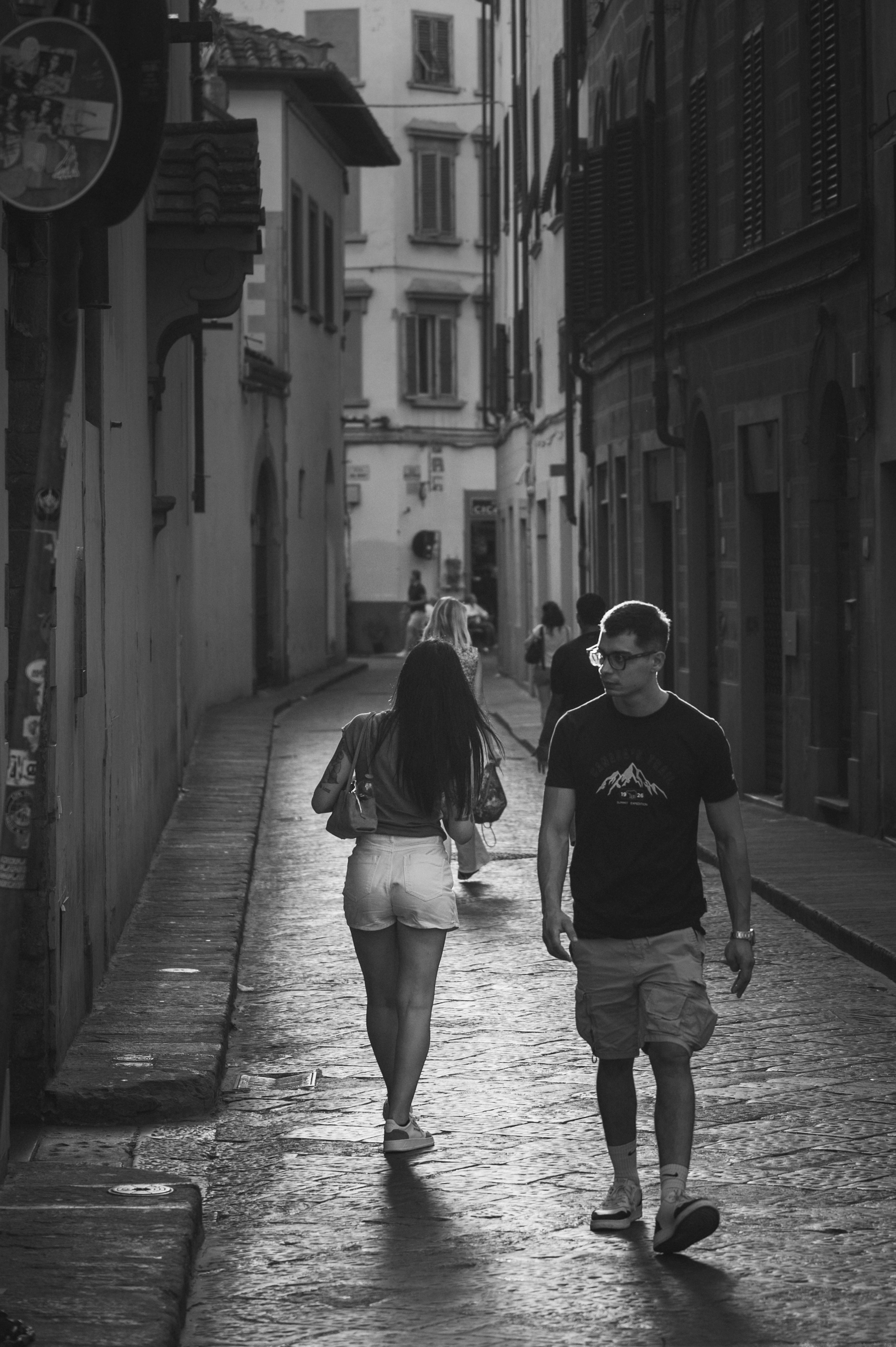 Monochrome street scene of pedestrians strolling along a narrow cobbled alley with sunlit reflections; the foreground figures draw the eye toward the distant pale façade. This is a photograph, capturing candid urban life.