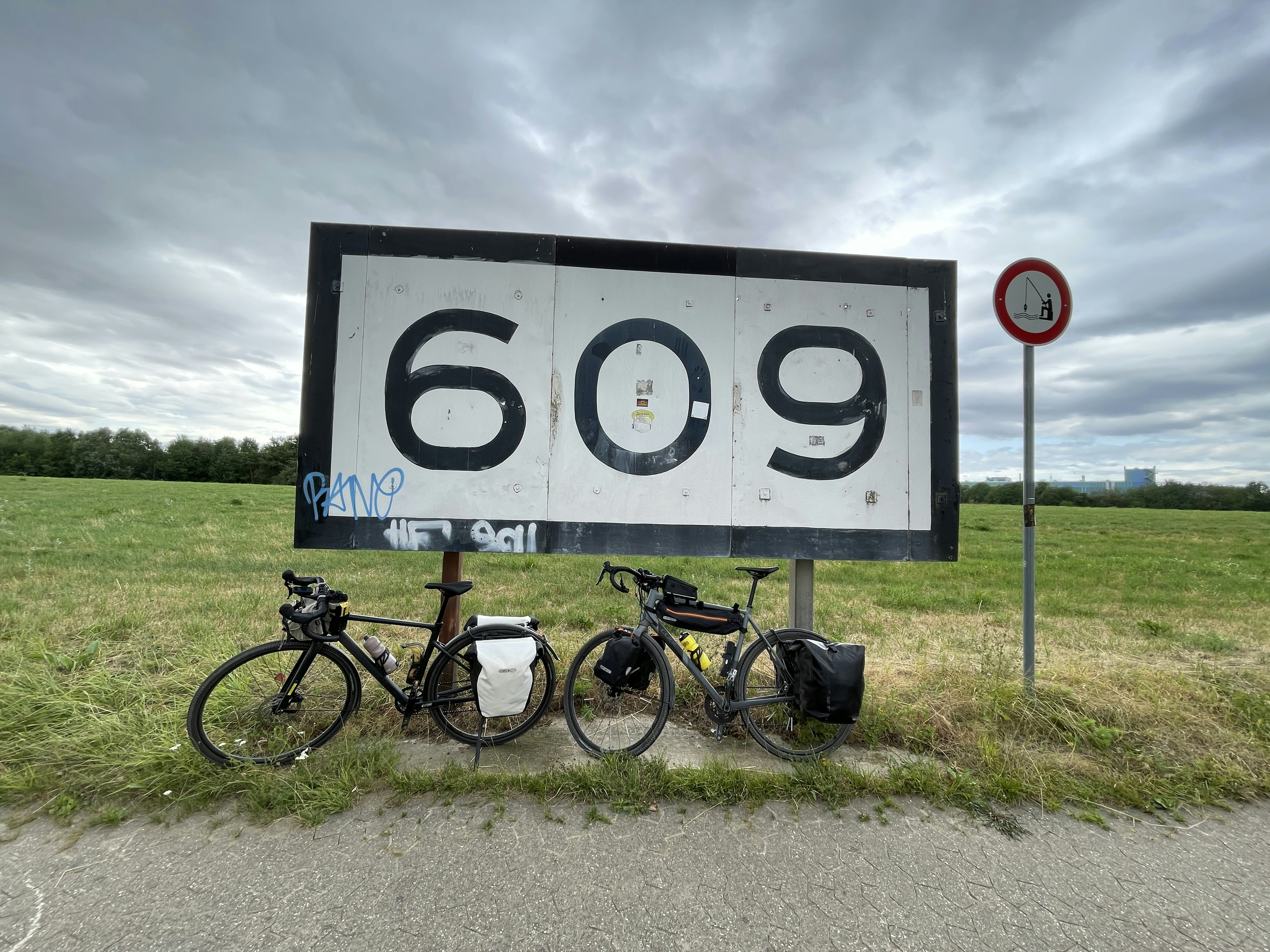 Two touring bicycles parked on a grassy verge beside a large sign displaying '609' with a lone circular sign nearby, under a cloudy sky in an expansive flat landscape.