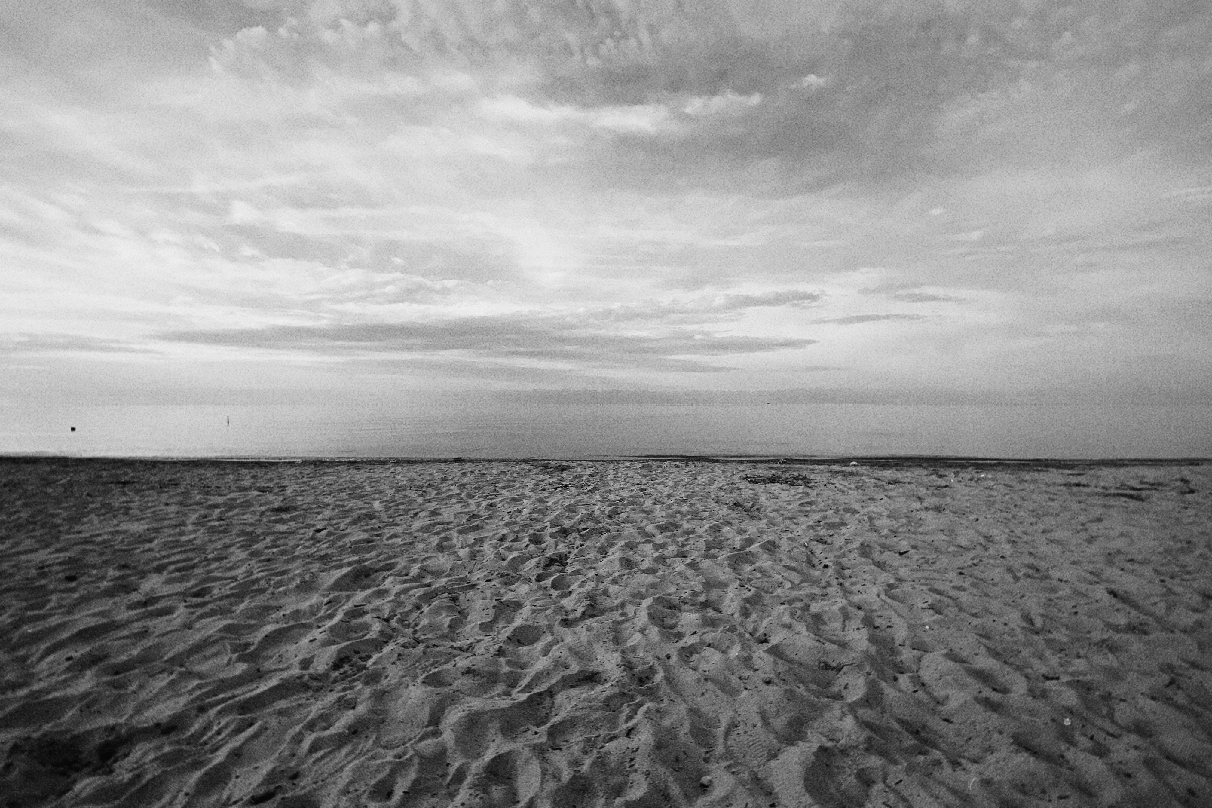 Low-contrast beach scene with a textured sandy foreground and distant calm horizon; monochrome rendering emphasizes pattern and mood without prominent figures or objects.